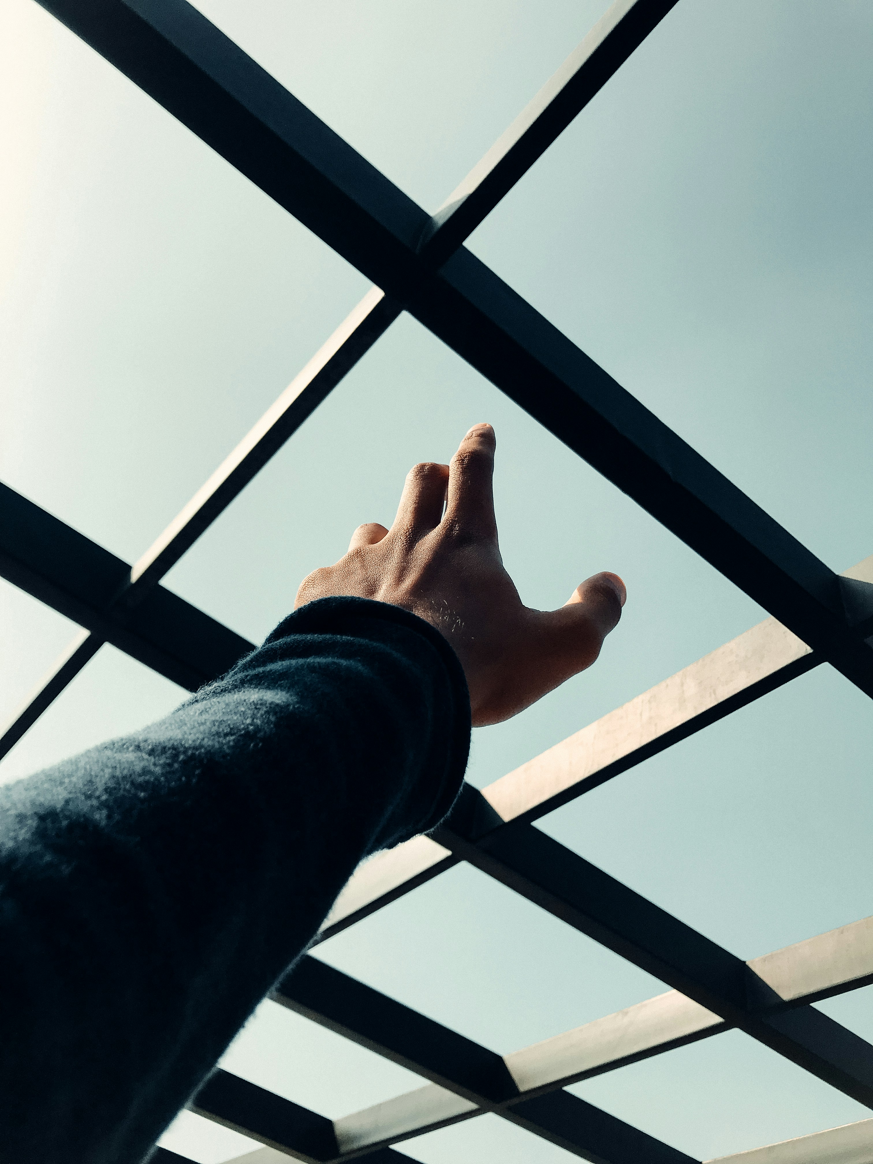 A hand extends towards a grid of dark beams against a pale sky, symbolizing aspiration and connection. The image captures a moment of longing and exploration.