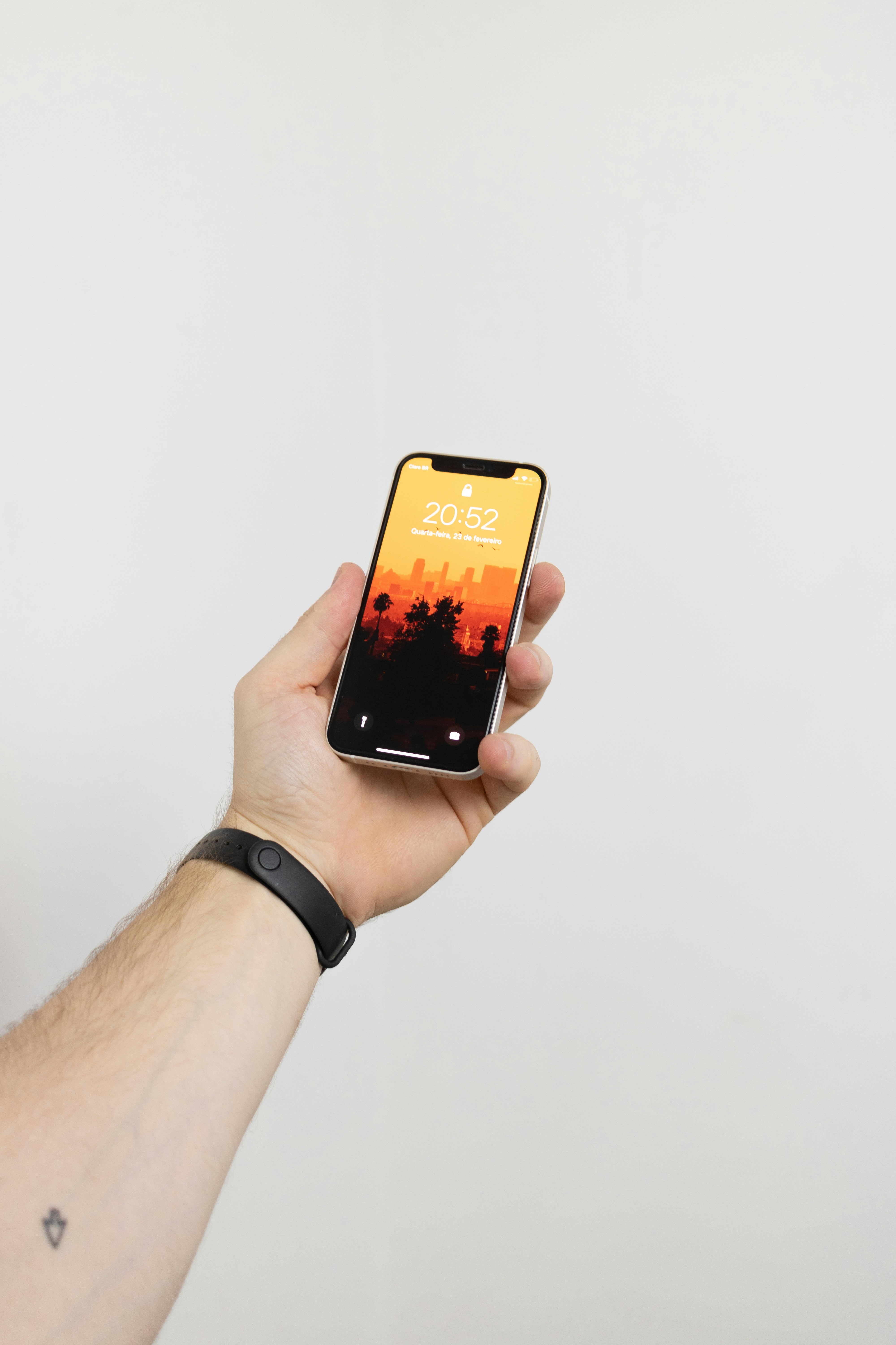 Hand holding a smartphone displaying a vibrant sunset scene over a city skyline. The background is a plain white wall.