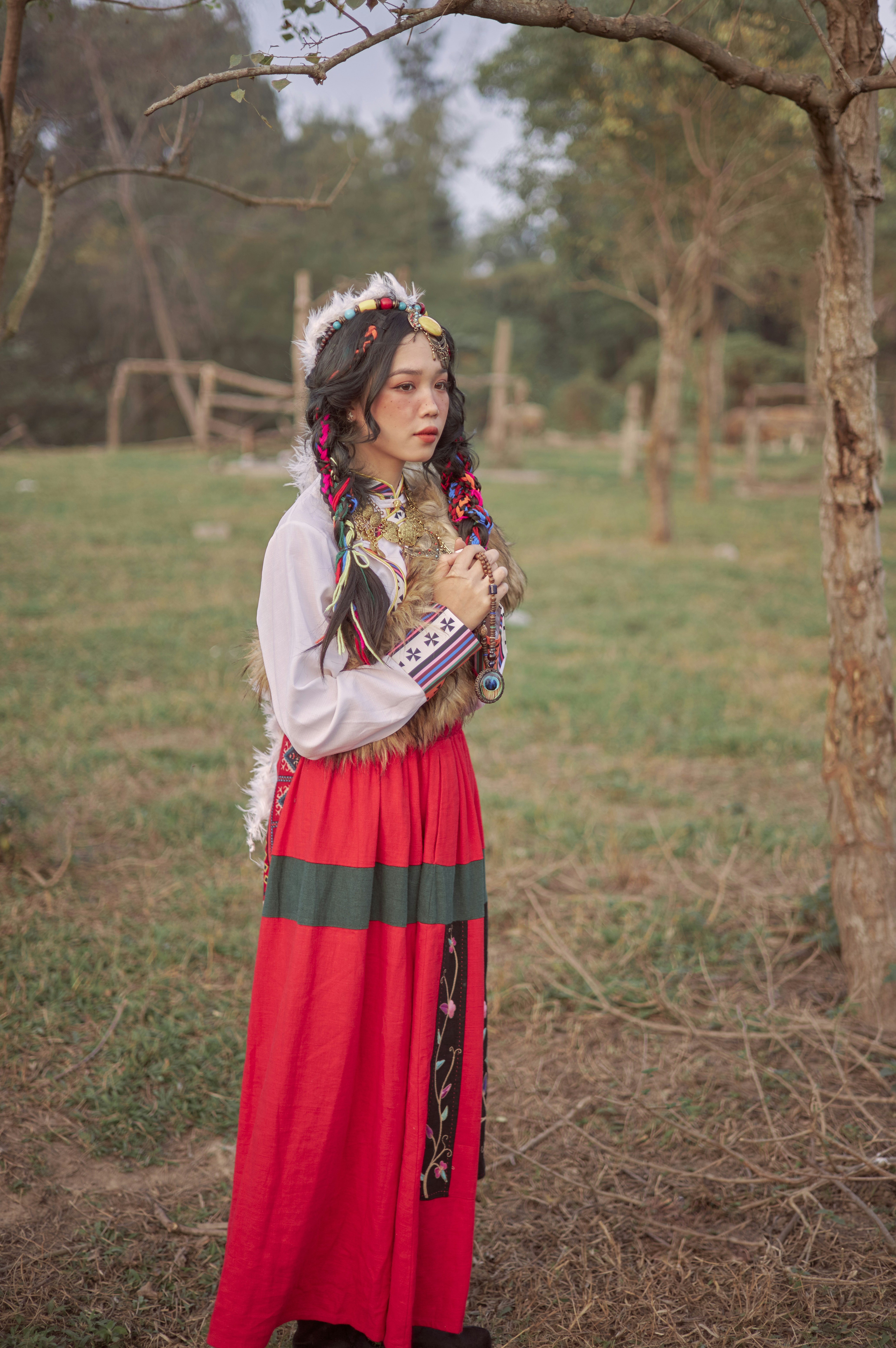 Young woman dressed in traditional attire, adorned with colorful accessories, standing in a serene outdoor setting.