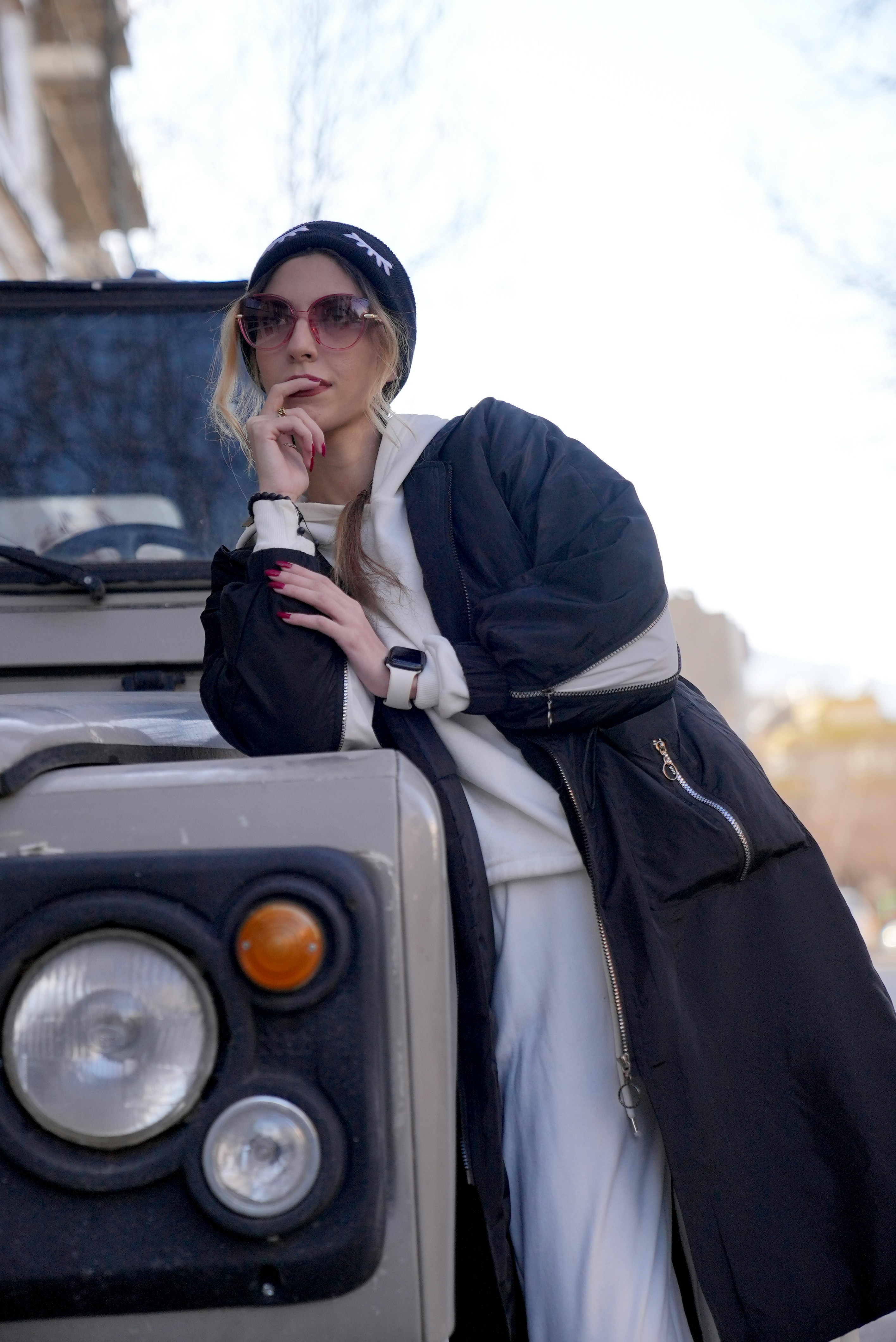 Urban cool: a woman leaning against a rugged SUV in a long black coat, hoodie, and sunglasses, posing with a thoughtful gaze. A logo-agnostic street portrait with a soft bokeh background.