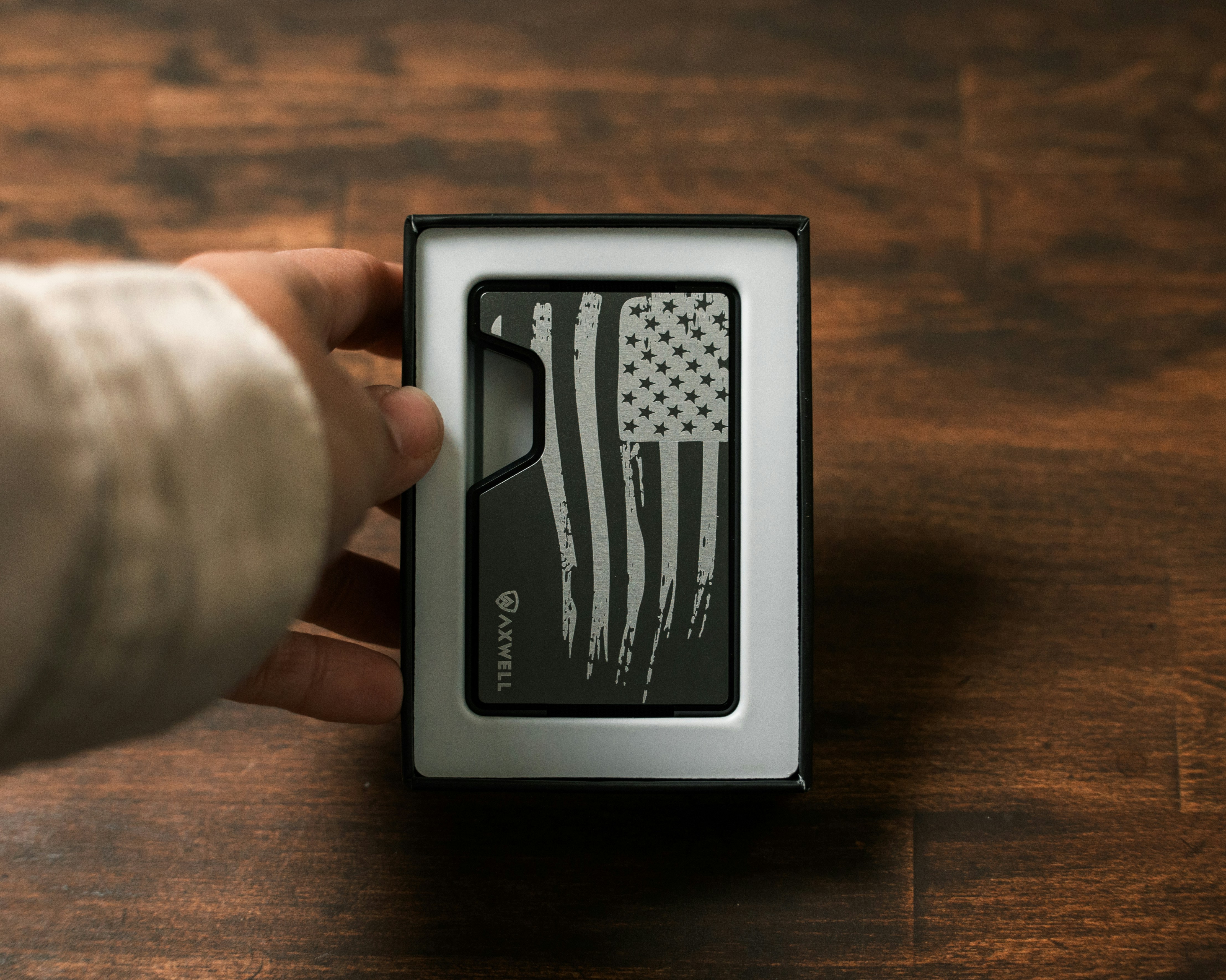 A hand holding a sleek wallet featuring a distressed American flag design, resting in its box on a wooden surface.