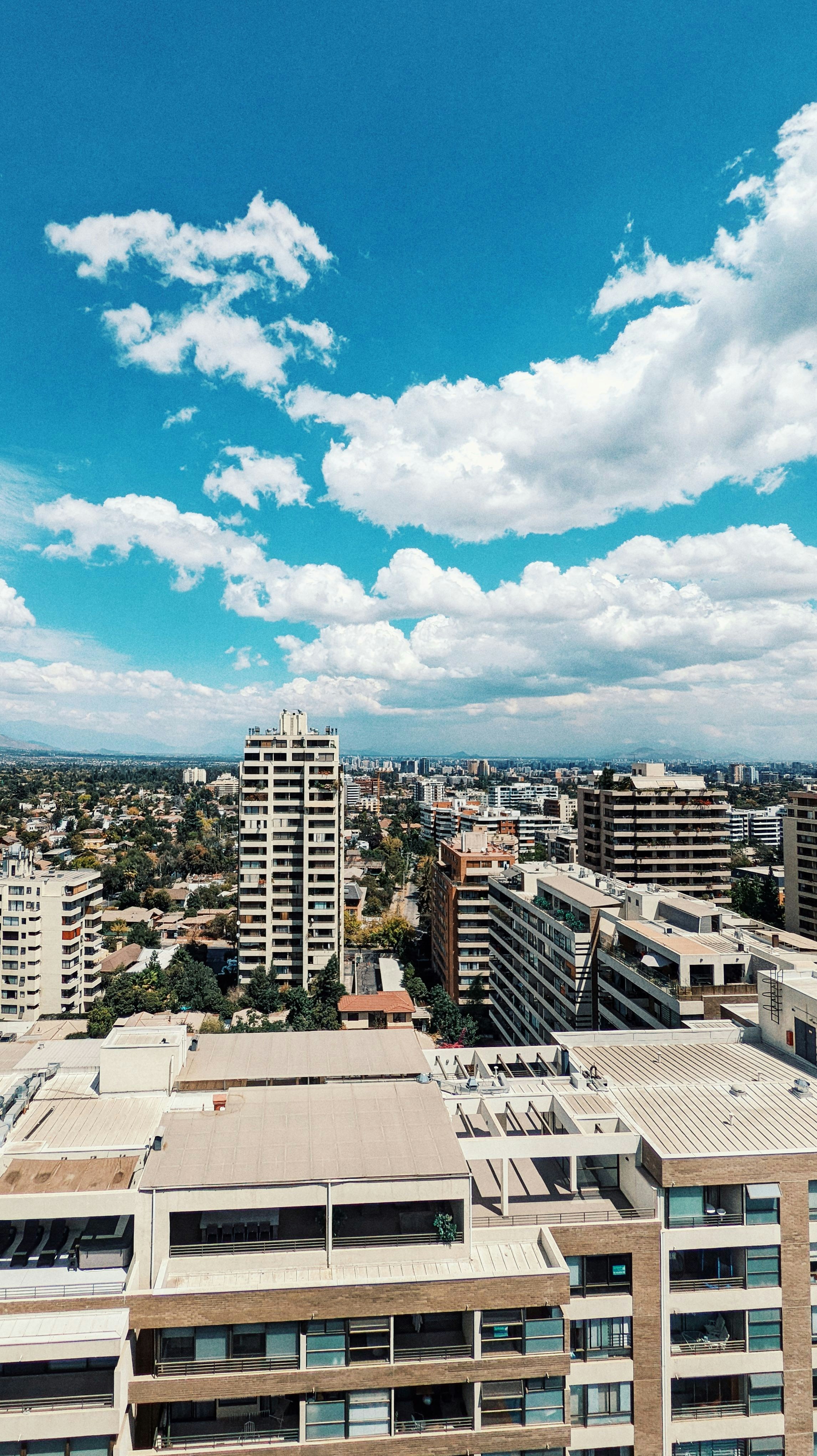 A panoramic view of a bustling cityscape featuring diverse architecture and lush greenery under a bright blue sky with fluffy clouds.