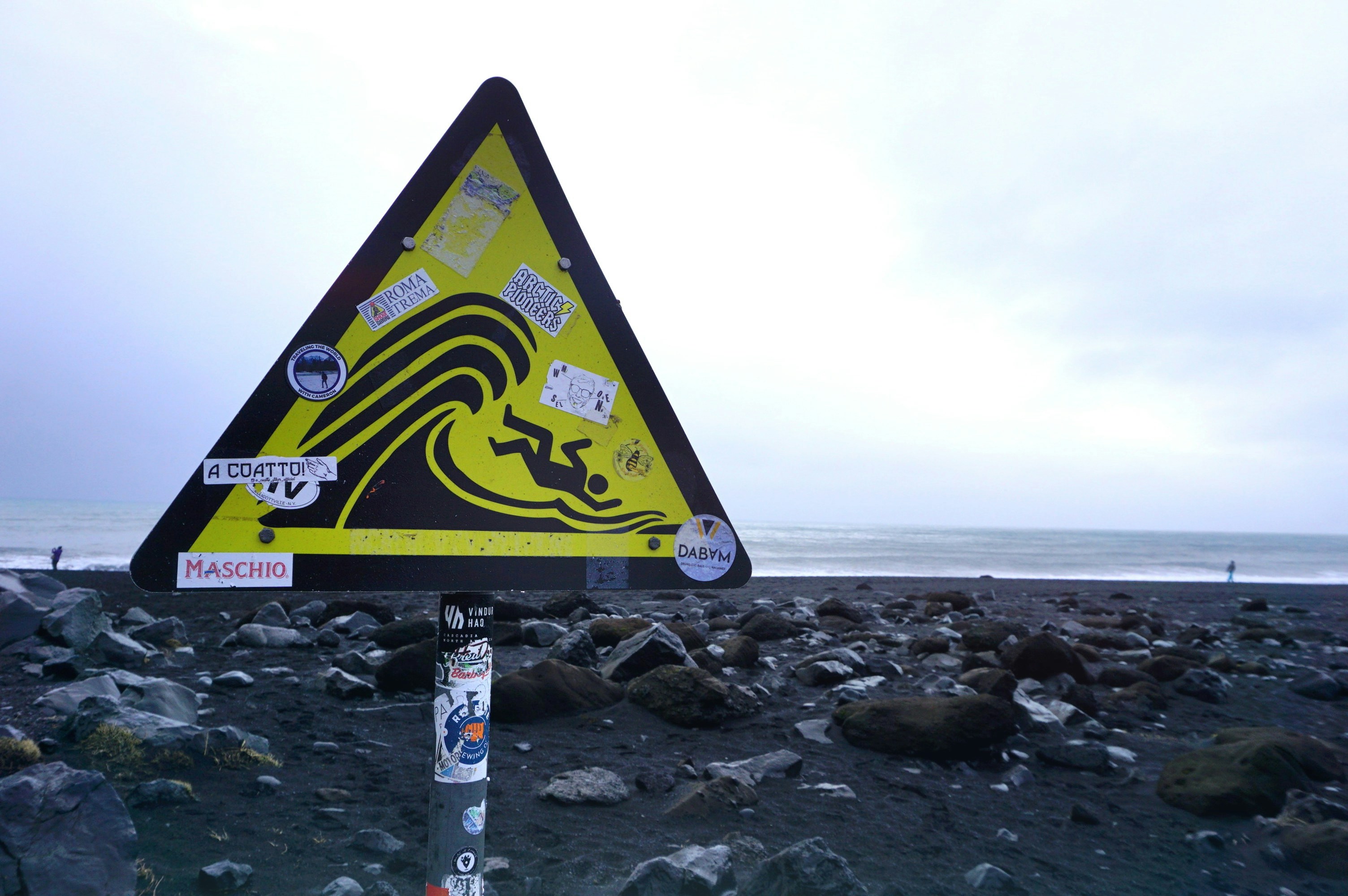 Vibrant yellow warning sign adorned with stickers, alerting beachgoers to potential waves, set against a rocky shoreline and gray ocean backdrop.