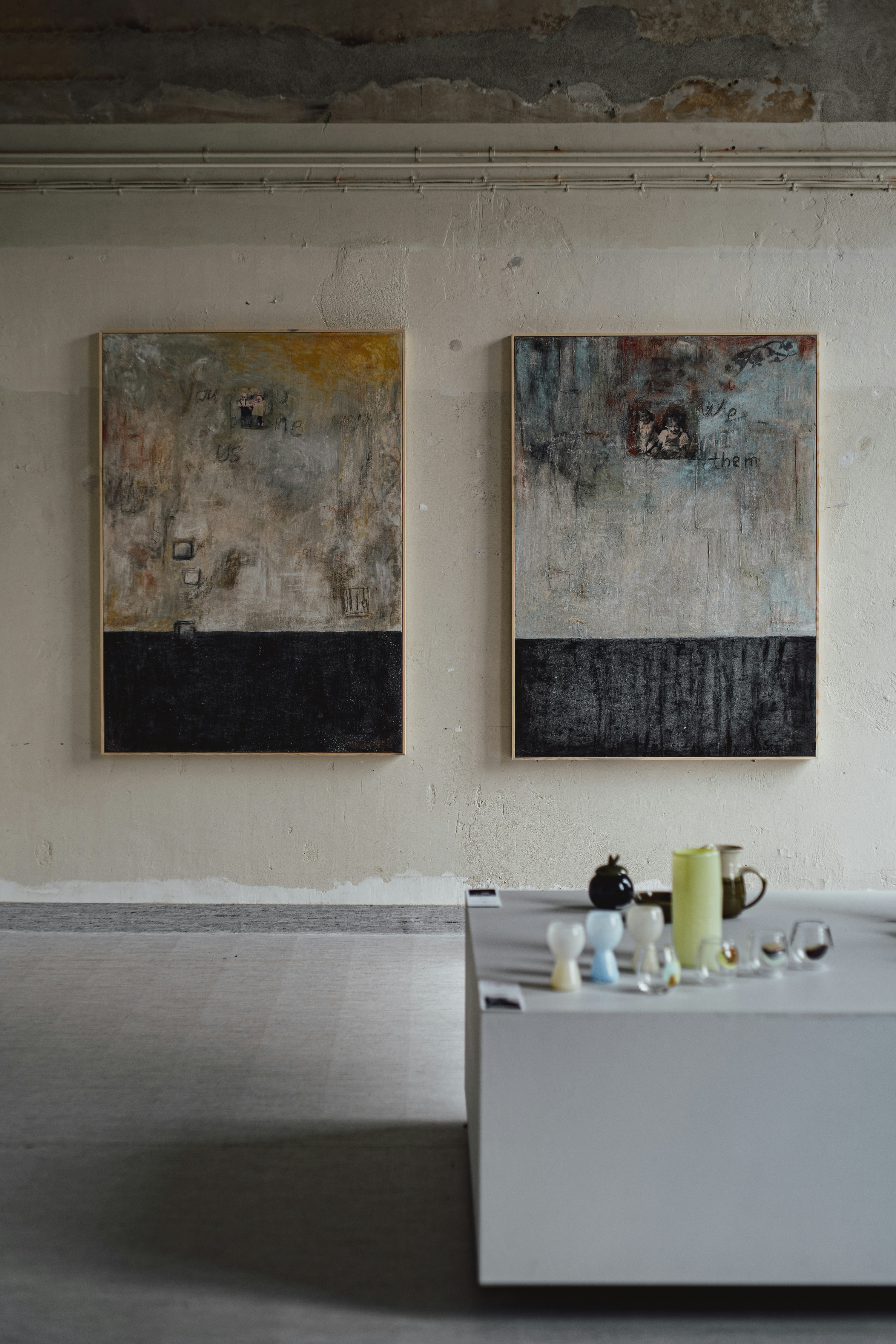 Two abstract paintings displayed on a textured wall, with a minimalist white table featuring various ceramic objects in the foreground.