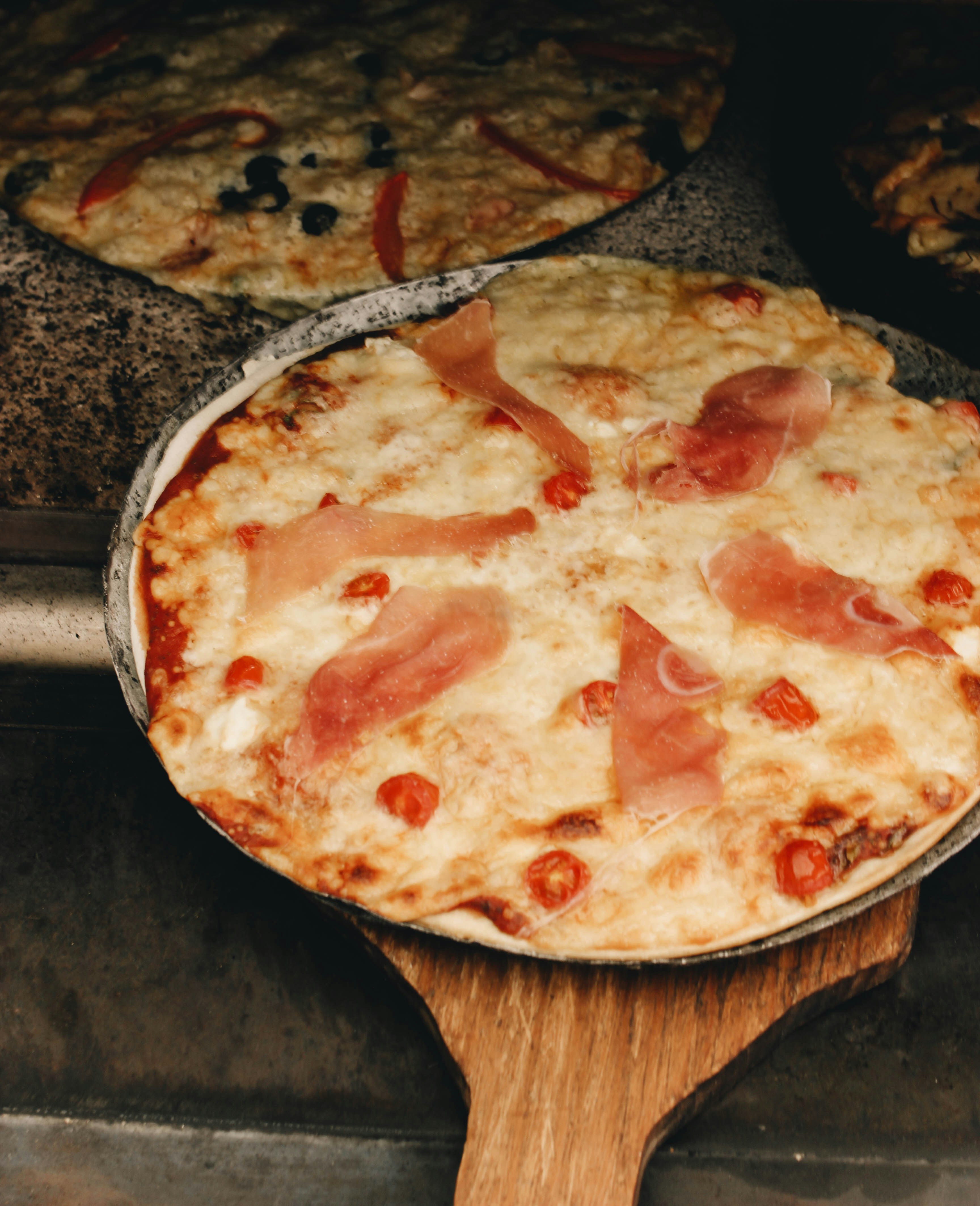 Freshly baked pizza topped with melted cheese, cherry tomatoes, and slices of ham, resting on a wooden peel in a rustic oven.