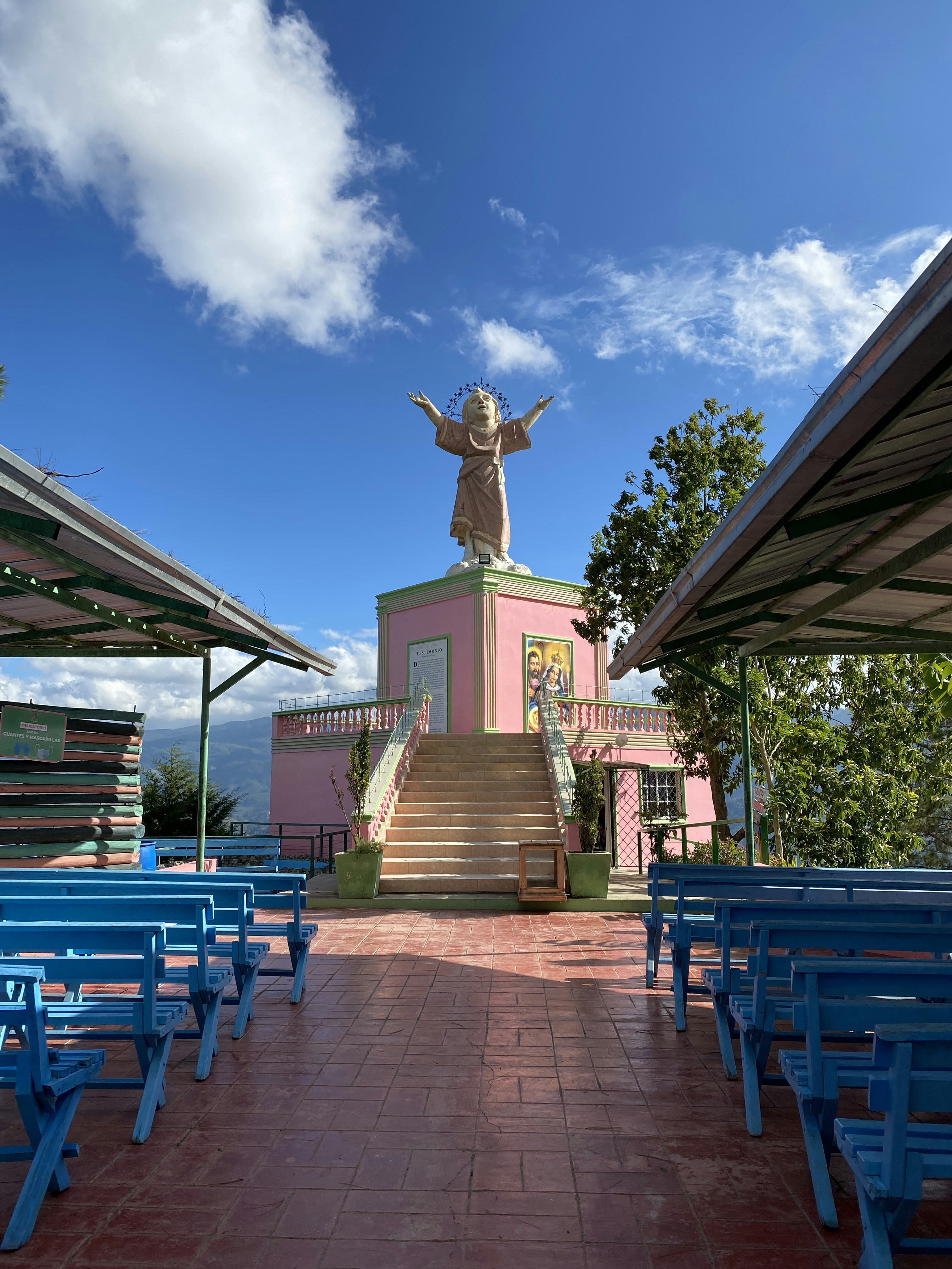 A grand statue with outstretched arms stands atop a pink platform, surrounded by blue benches and lush greenery under a vibrant sky.