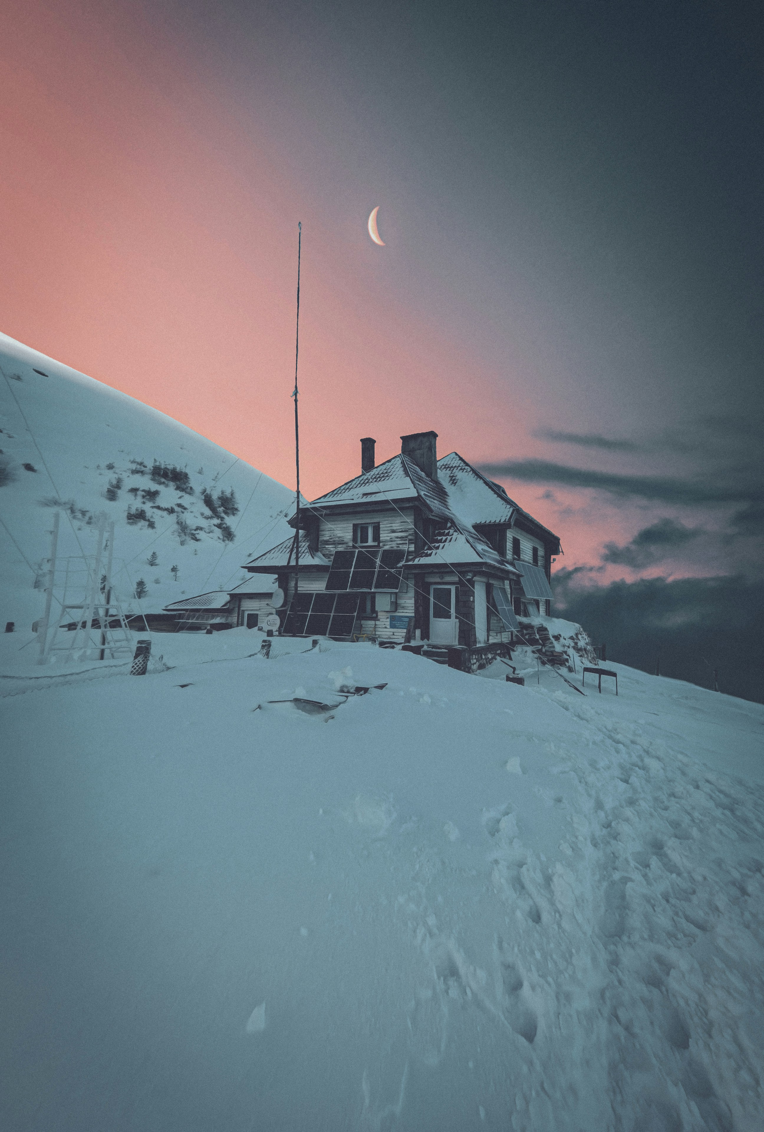 A solitary mountain cabin stands resilient against a backdrop of twilight hues, with a crescent moon illuminating the snowy landscape.