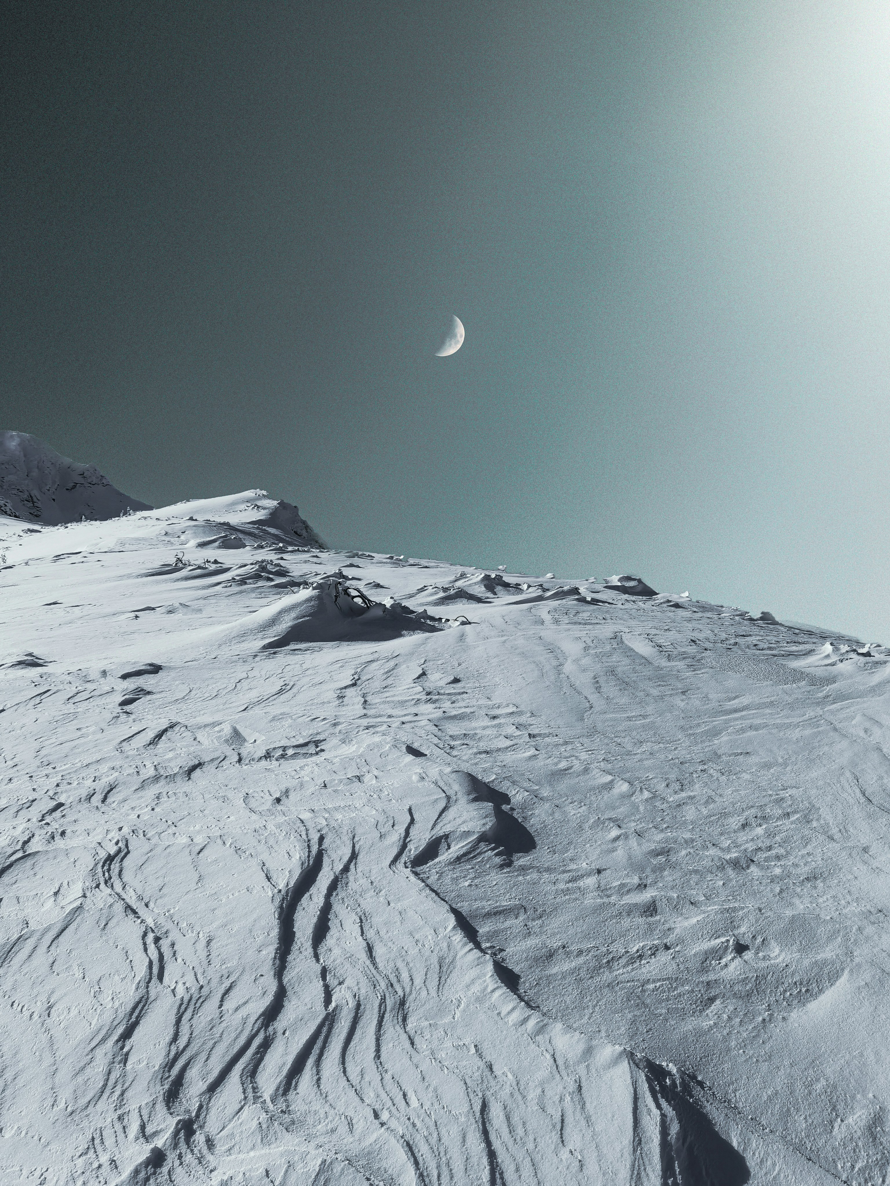 Crescent moon above a wind-sculpted snowfield with distant ridges under a pale blue sky.