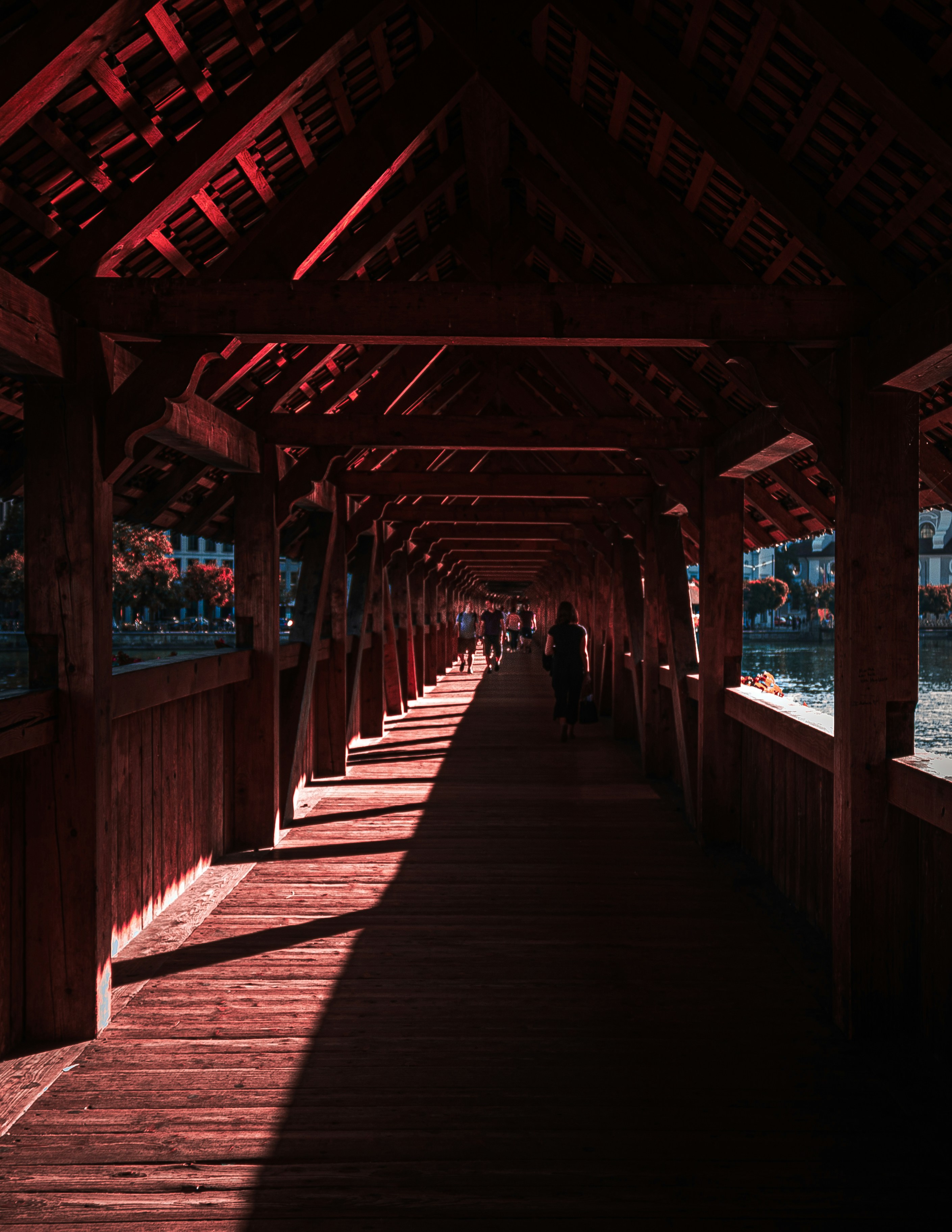 Wooden walkway beneath a slatted roof, illuminated by warm light and deep shadows, leading toward distant figures by the water's edge.