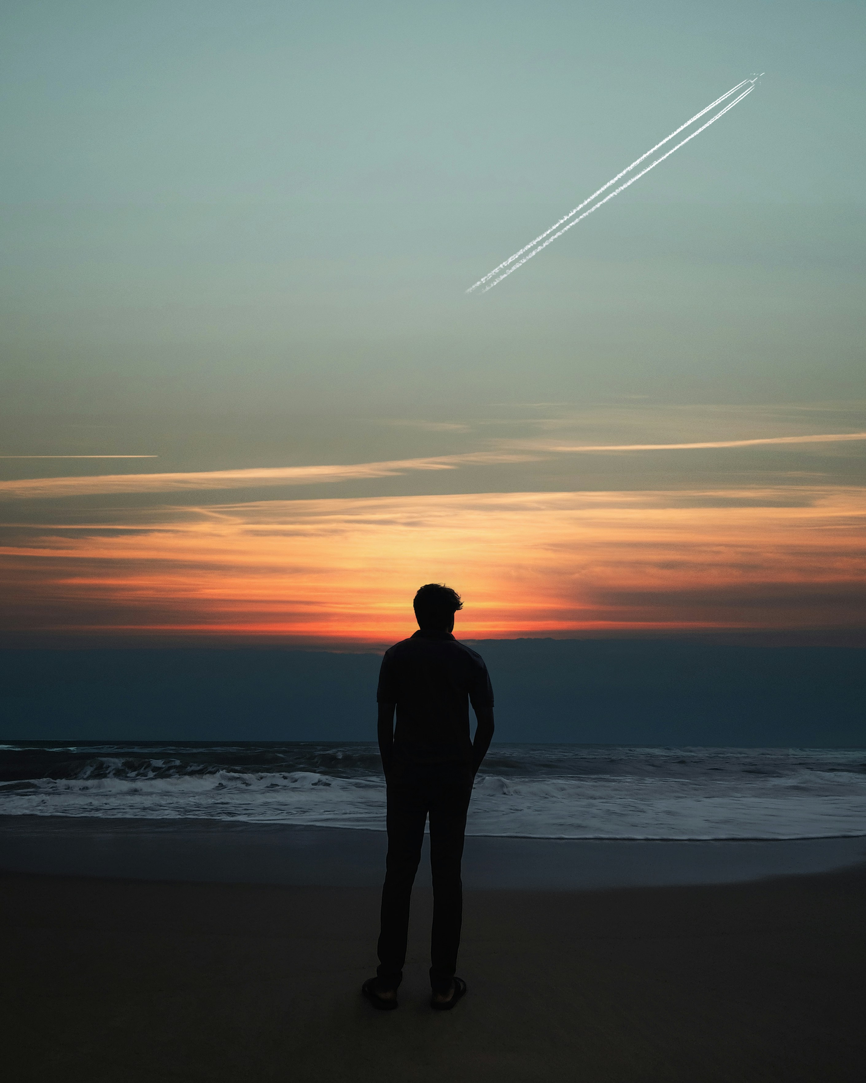 Silhouette of a person standing on the beach, gazing at the sunset with airplane contrails streaking the sky.