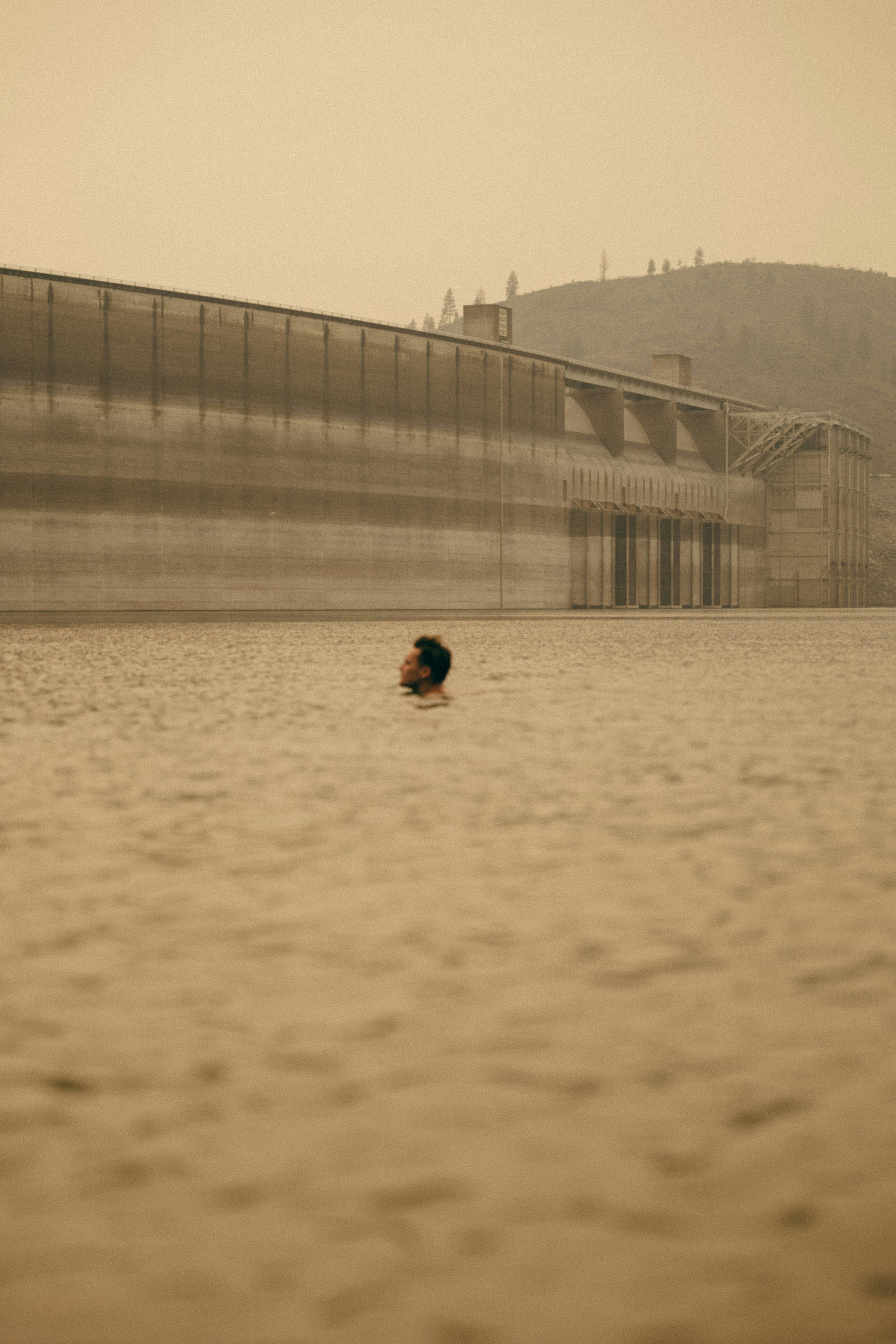 A solitary figure wades through murky waters under a smoky sky, with a distant industrial structure looming in the background.