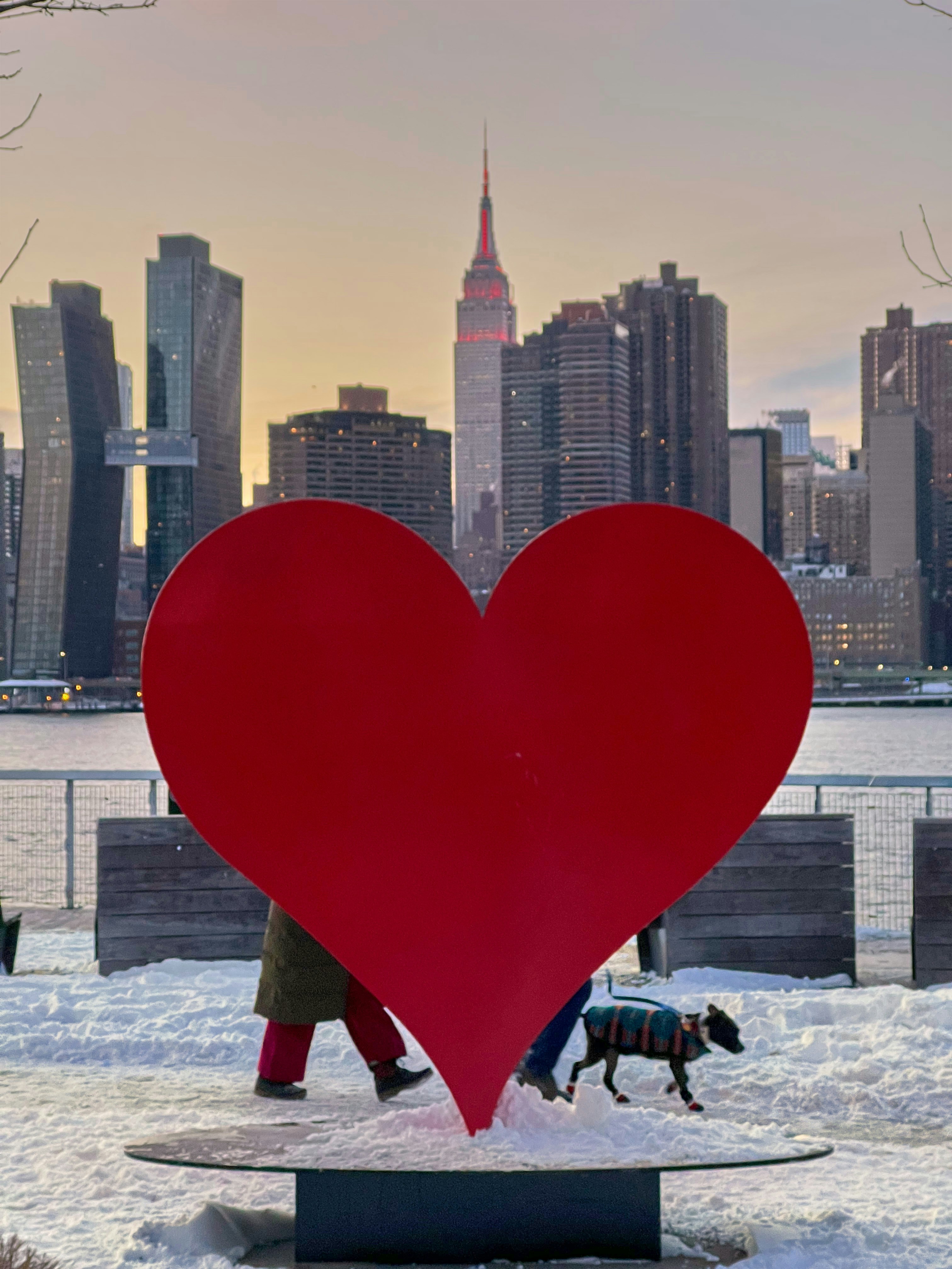 Red heart sculpture in snow with the Empire State Building illuminated in red at sunset.