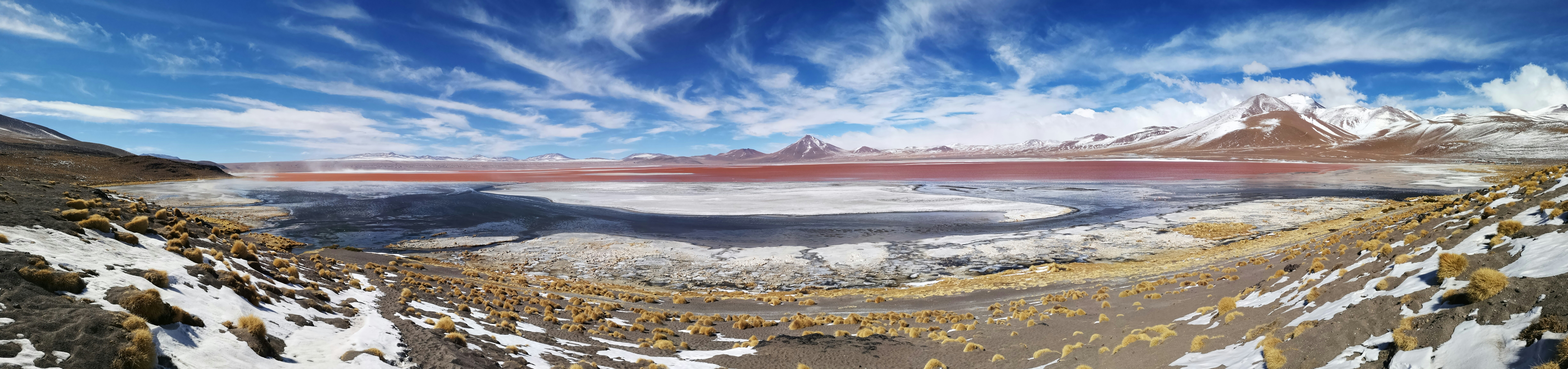 Vast panoramic view of the Altiplano landscape, showcasing a vibrant red lake surrounded by snow-capped mountains and a dramatic sky.