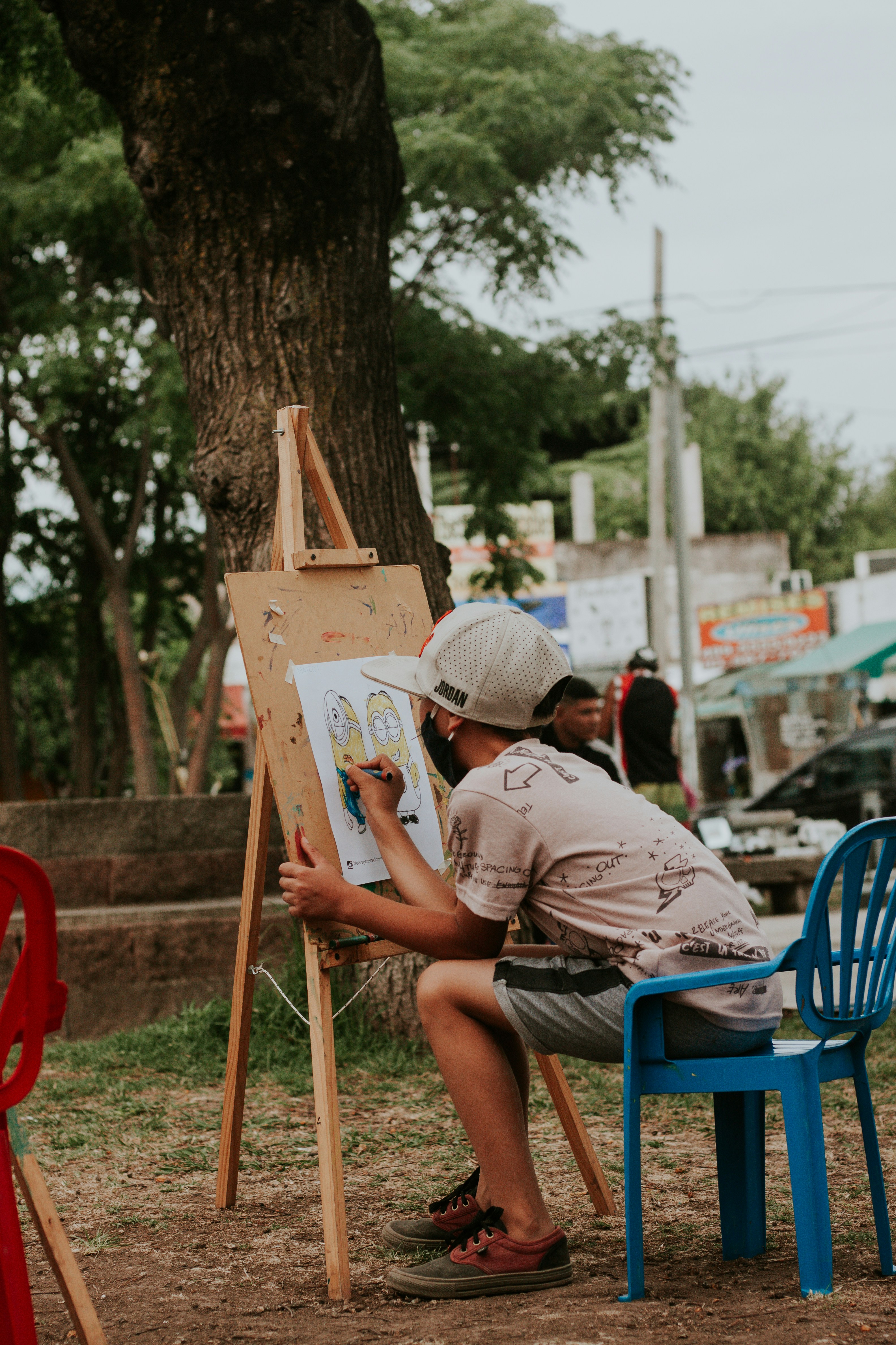 Child drawing animated characters on an easel in a lively outdoor park. Surrounding greenery and casual seating enhance the creative atmosphere.