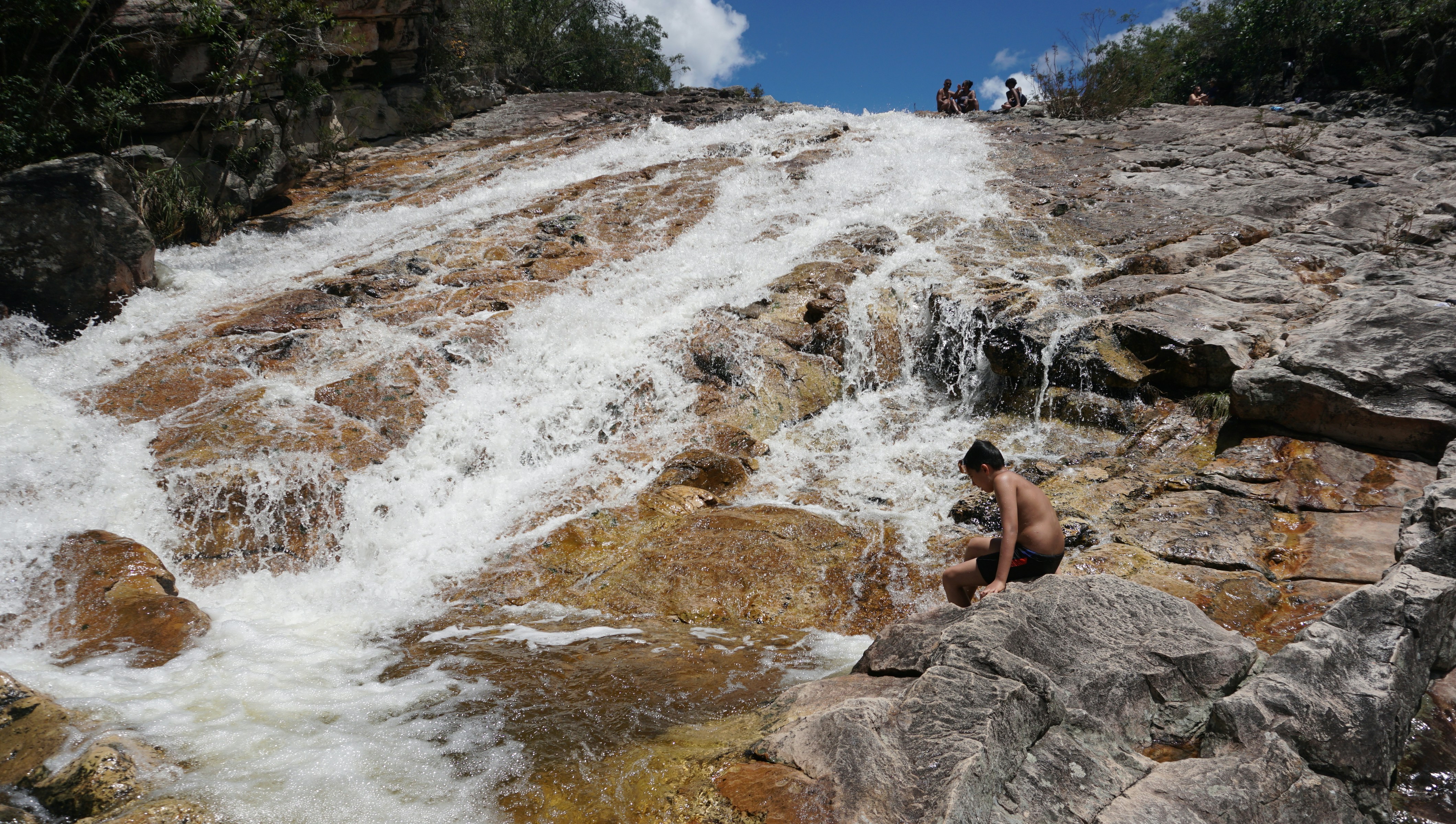 Person sitting beside a cascading waterfall over rocky terrain under a clear blue sky.