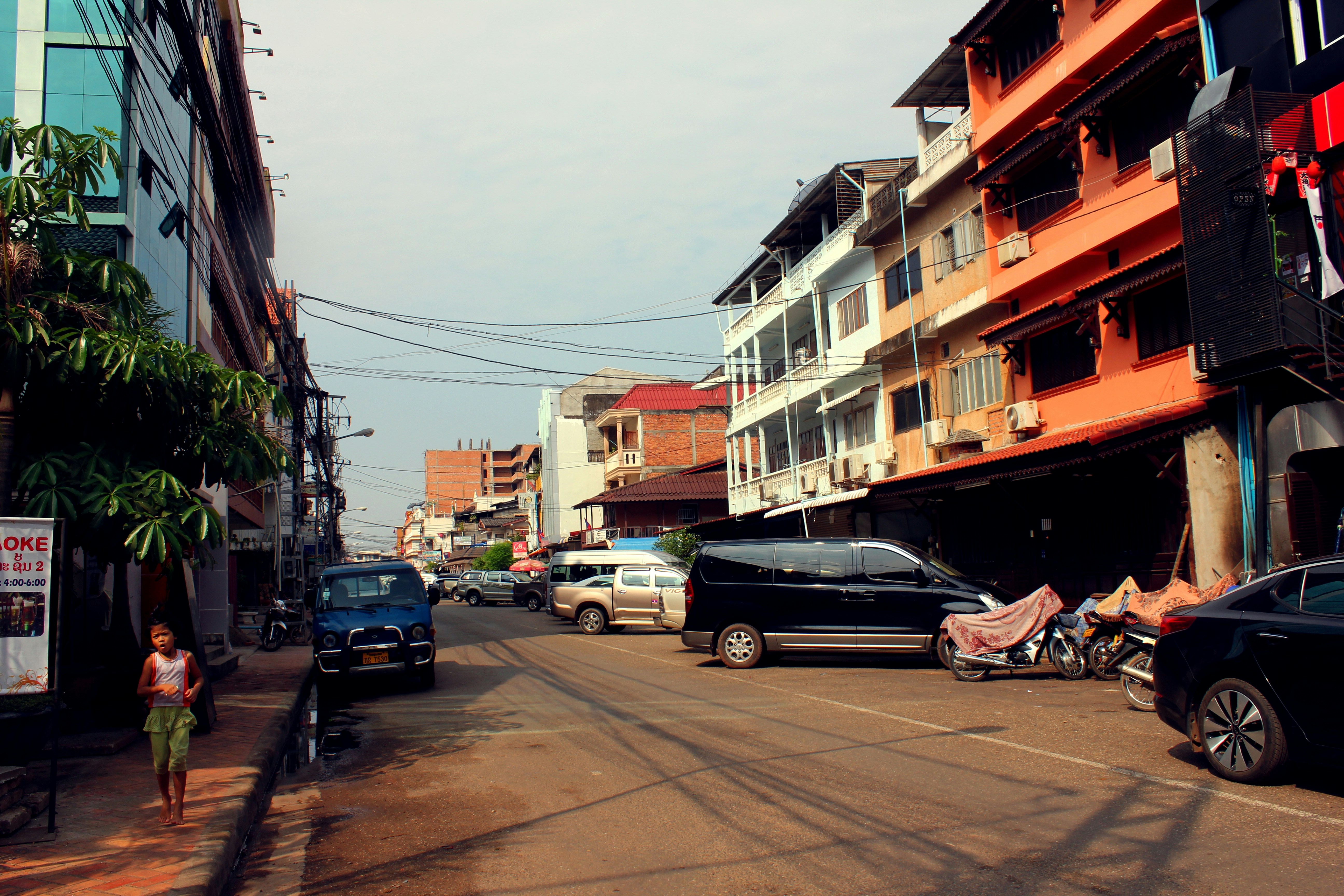 A bustling street scene showcasing a mix of parked vehicles and local architecture, with a child walking along the sidewalk. The urban landscape reflects daily life in a vibrant community.