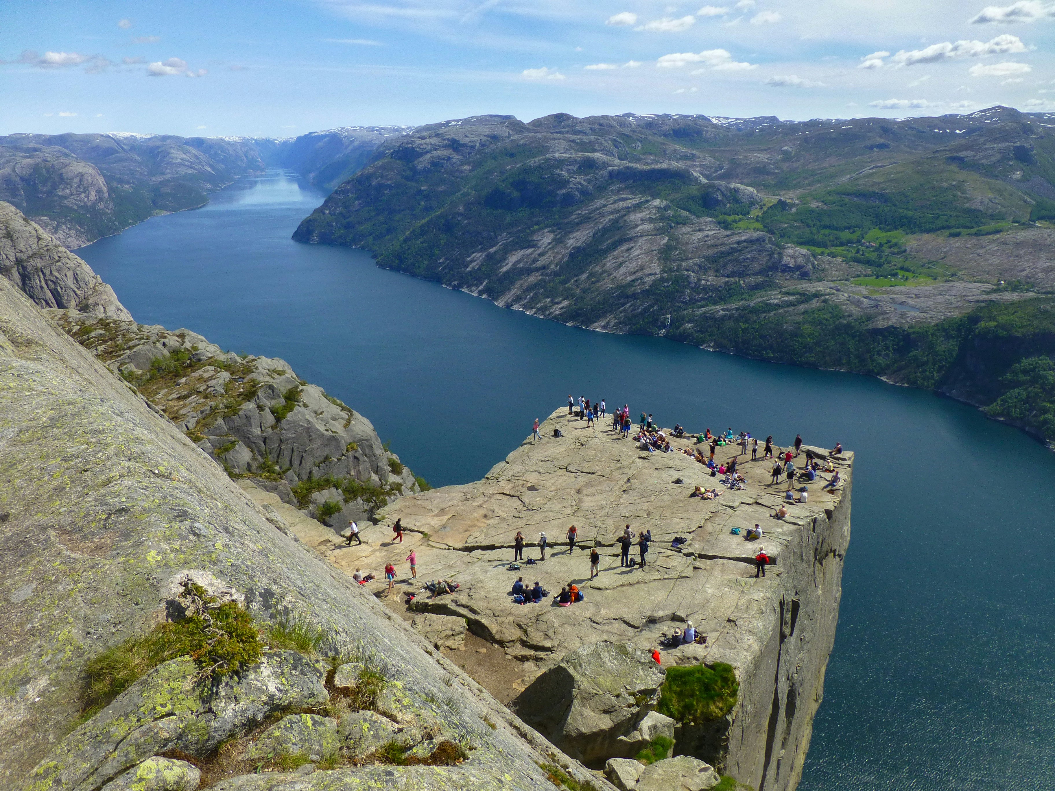 Visitors gather on the iconic cliff edge overlooking a deep fjord surrounded by rugged mountains and lush greenery.
