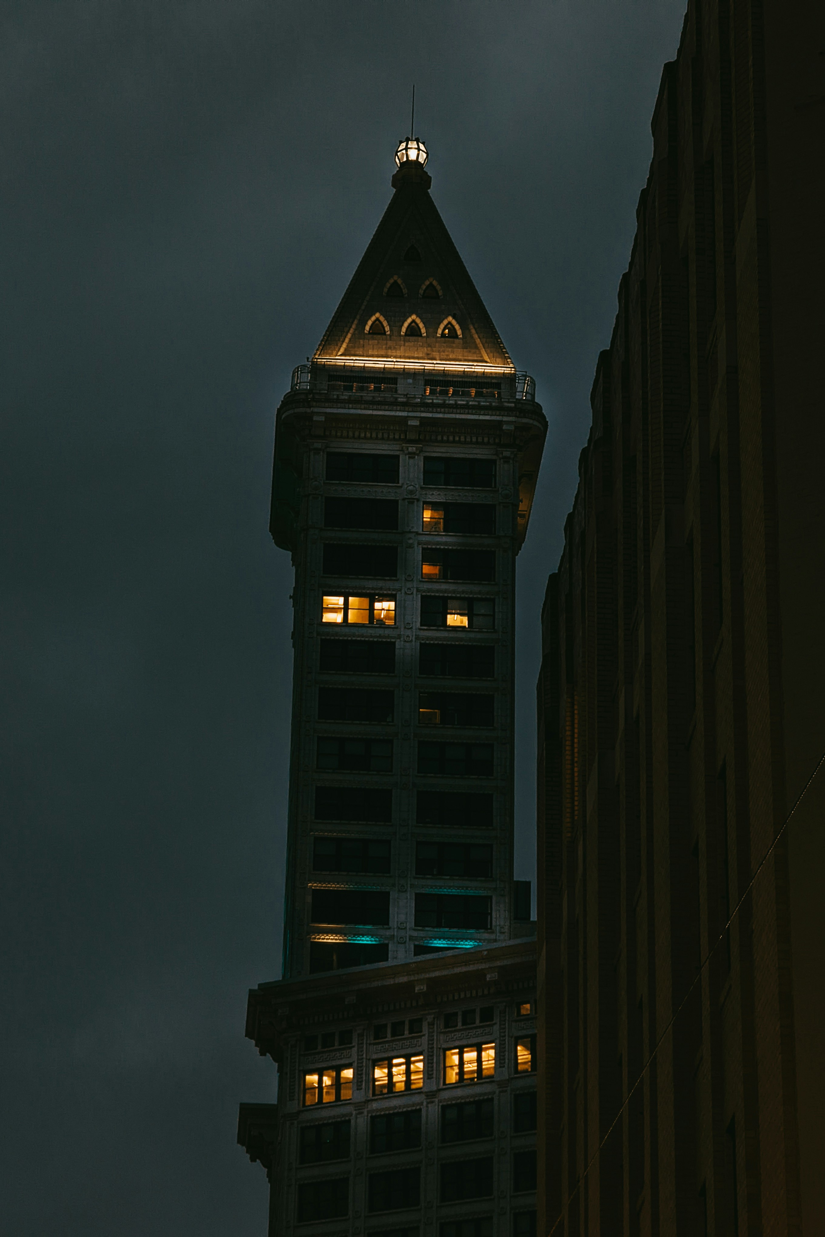 Night photograph of a tall skyscraper with an illuminated pyramidal crown rising beside a darker facade against a moody, cloudy sky. Warm window lights behind the grid of windows hint at occupancy.