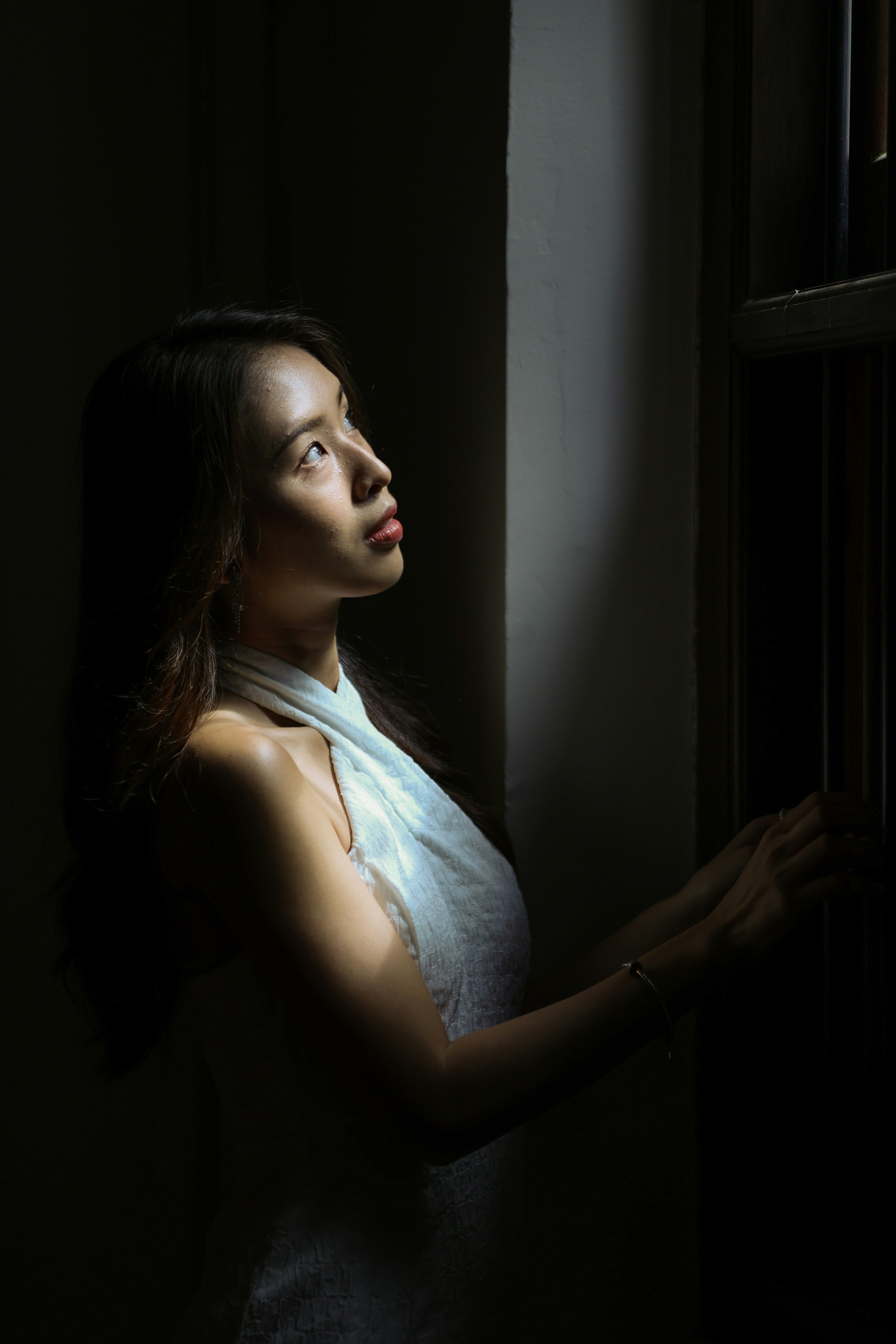 A woman gazes thoughtfully towards a window, illuminated by a soft beam of light that highlights her features against a dark background.