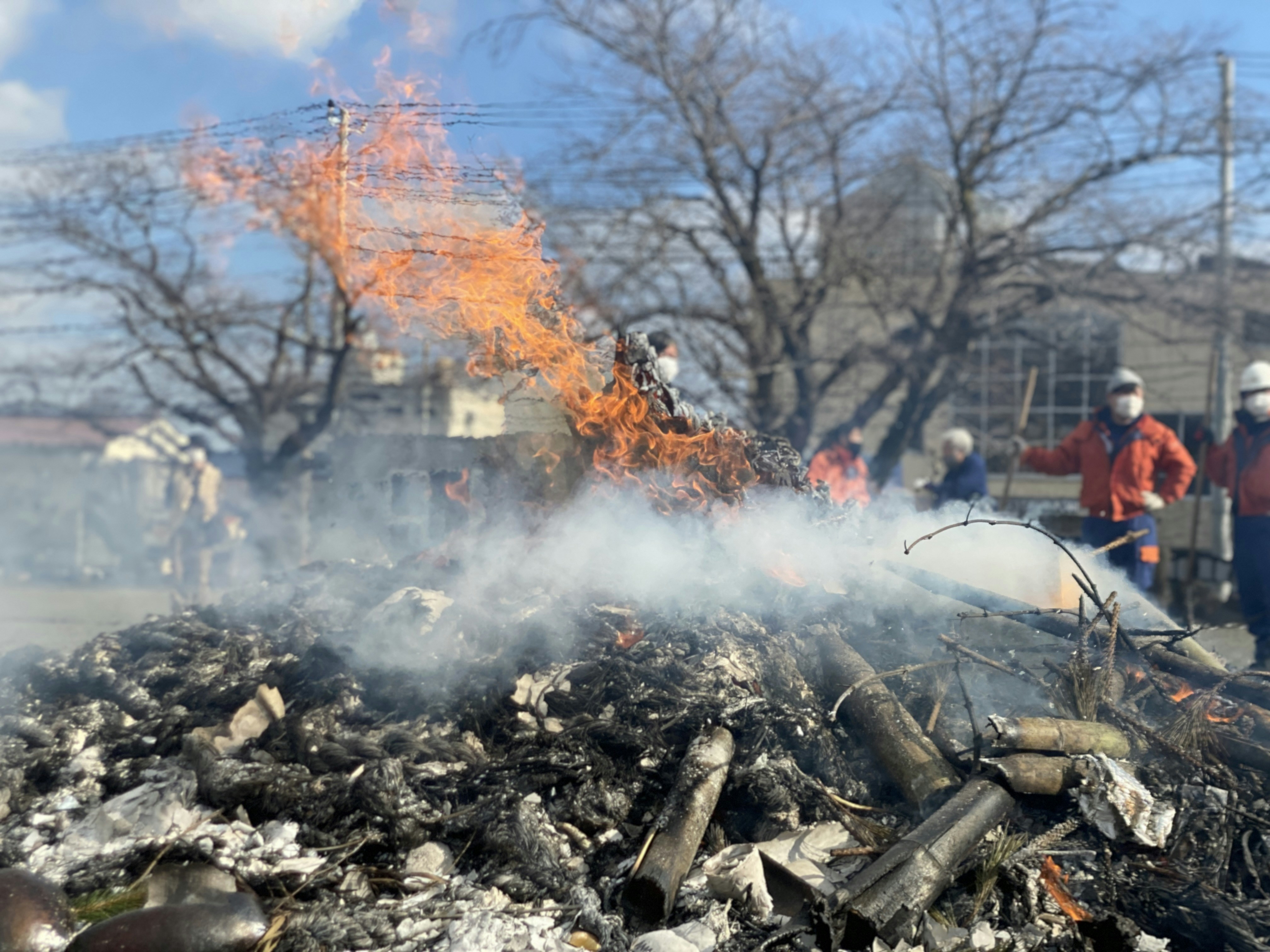 Vivid flames rise from a smoldering pile of wood with onlookers in the background, framed by bare winter trees.