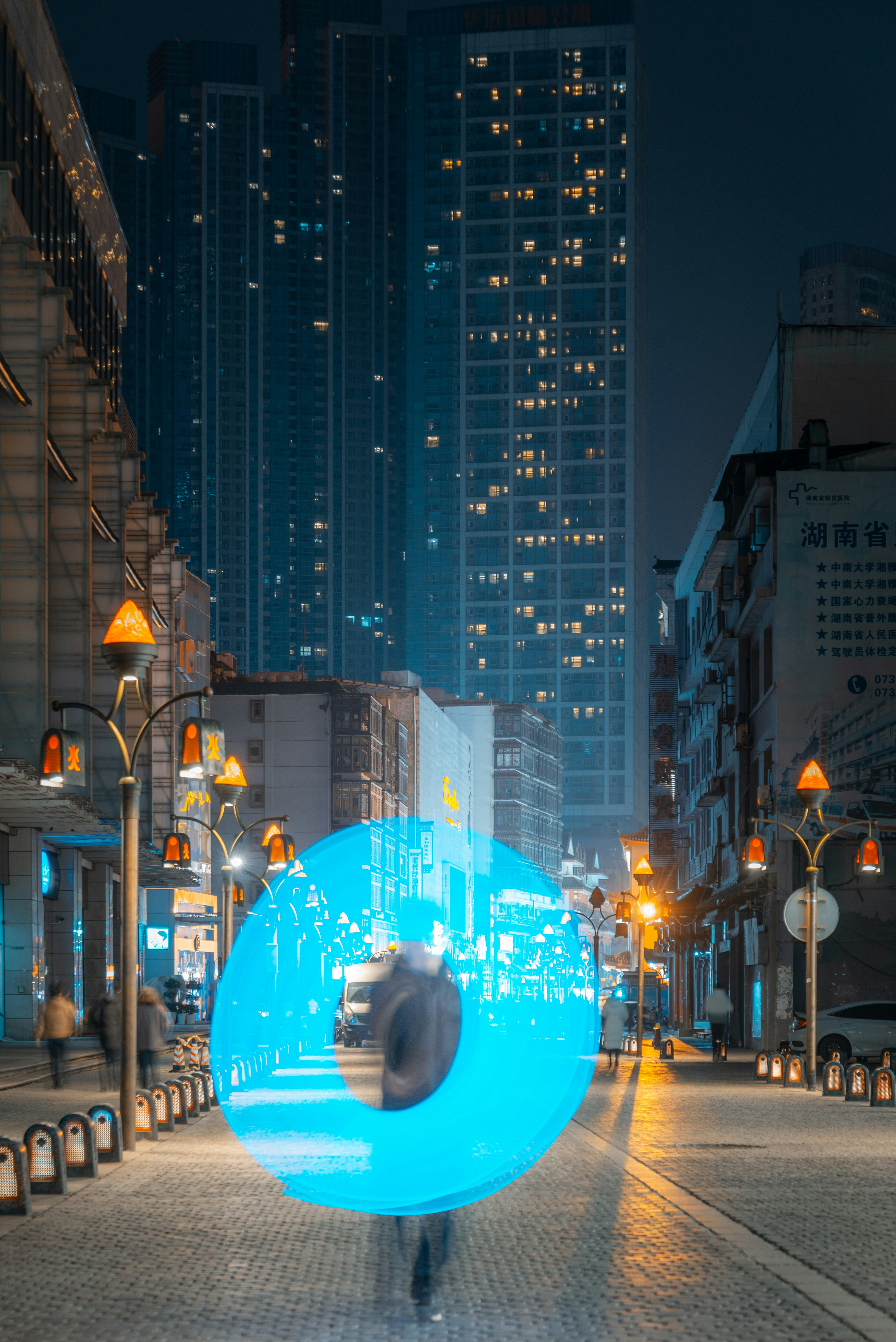 A blurred figure moves through a vibrant urban street, illuminated by glowing blue light, surrounded by towering skyscrapers and warm street lamps.