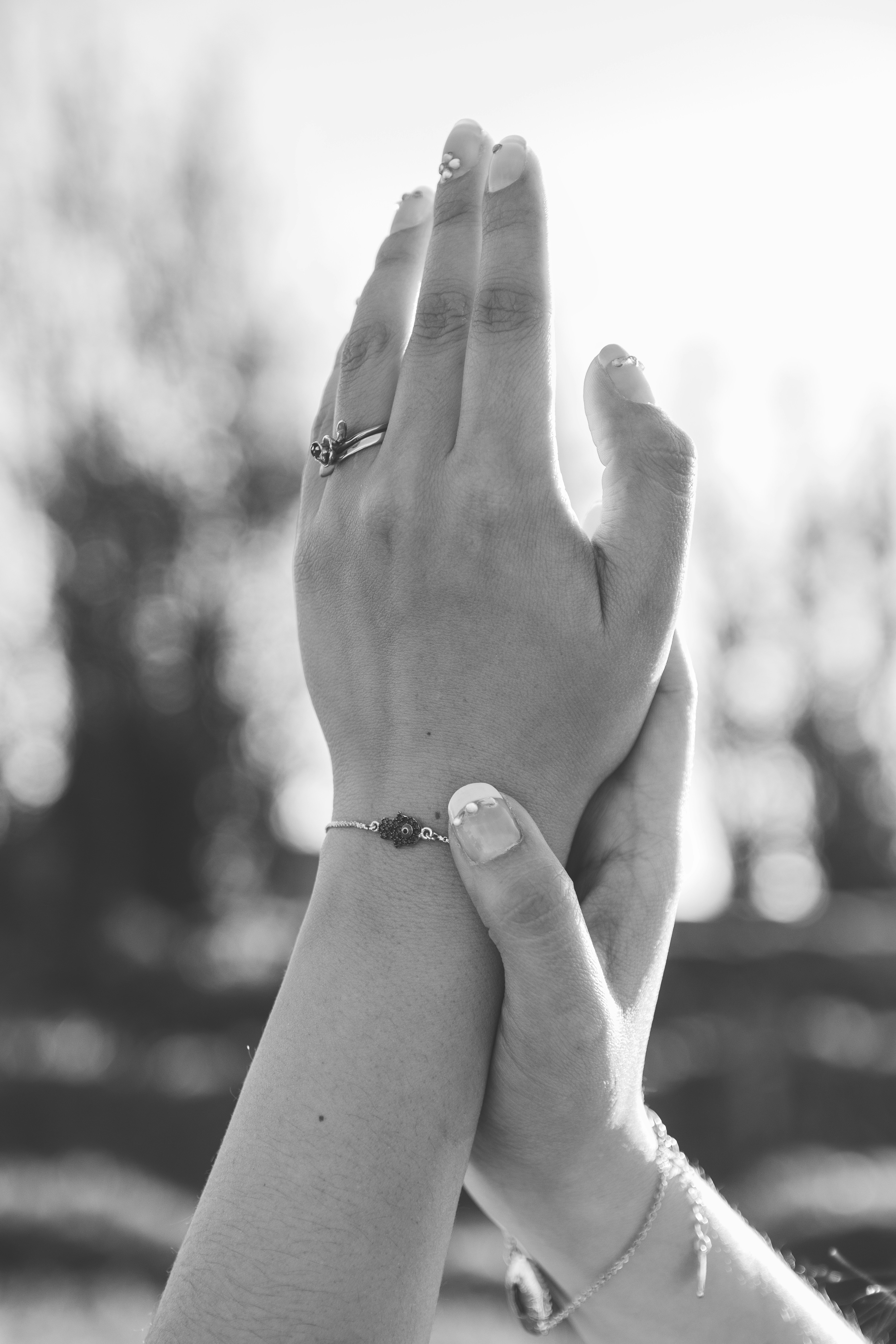 Two hands intertwined, showcasing delicate jewelry against a blurred natural backdrop. A moment of unity captured in monochrome.