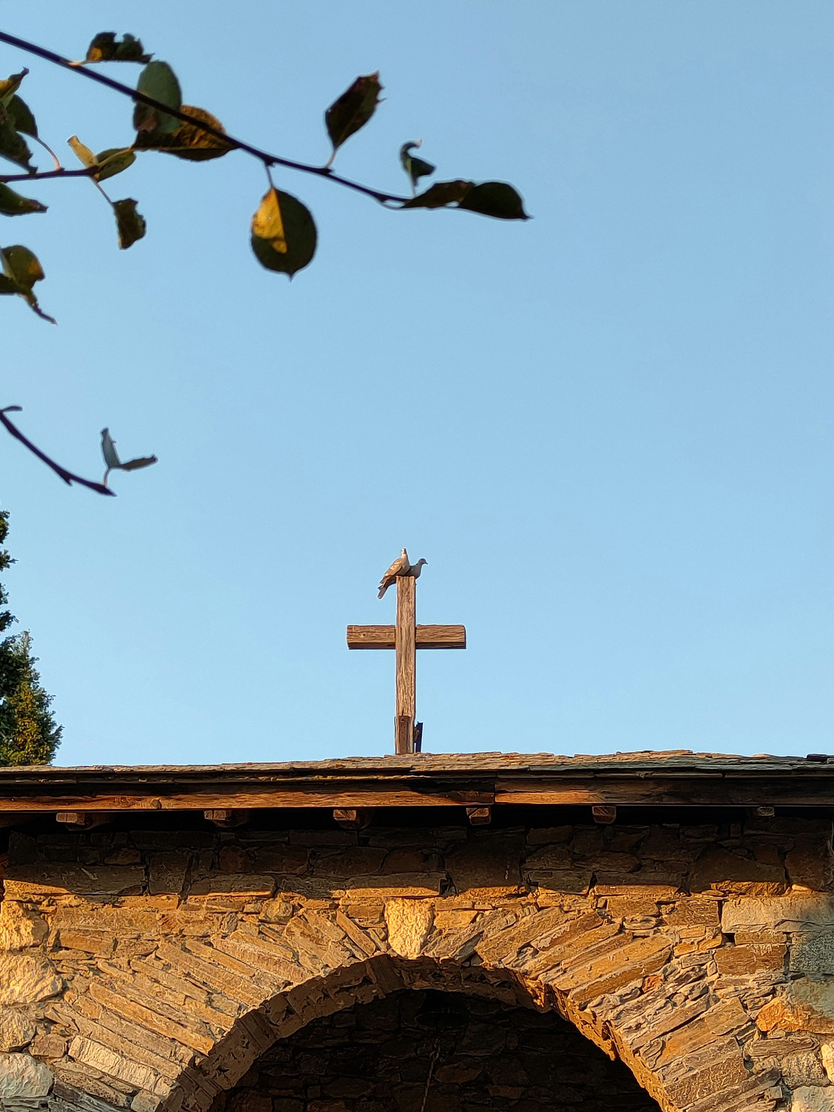 Wooden cross atop a rustic stone structure framed by leaves under a clear blue sky.
