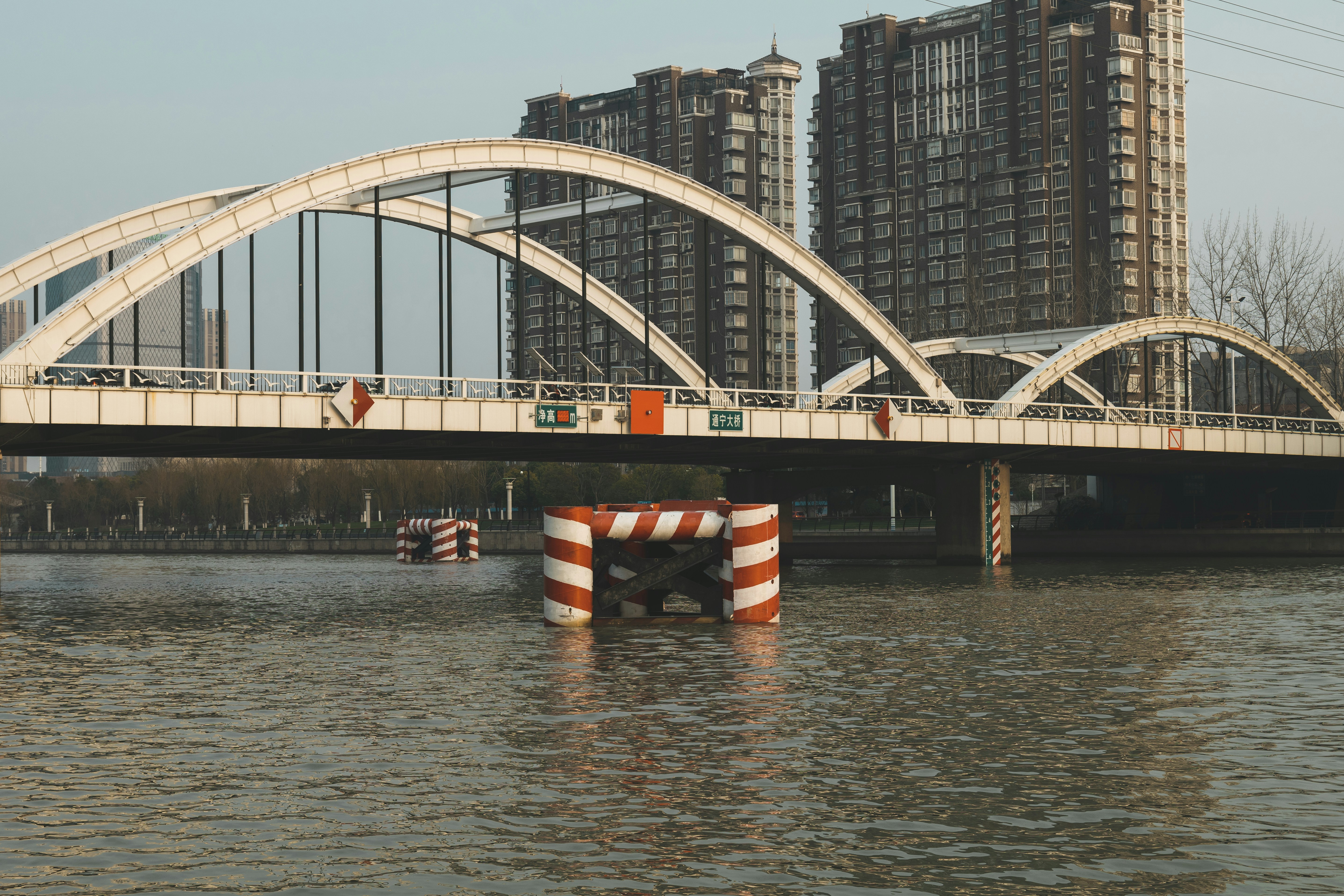 A modern bridge arches gracefully over a calm river, framed by urban buildings, with safety markers visible in the water below.