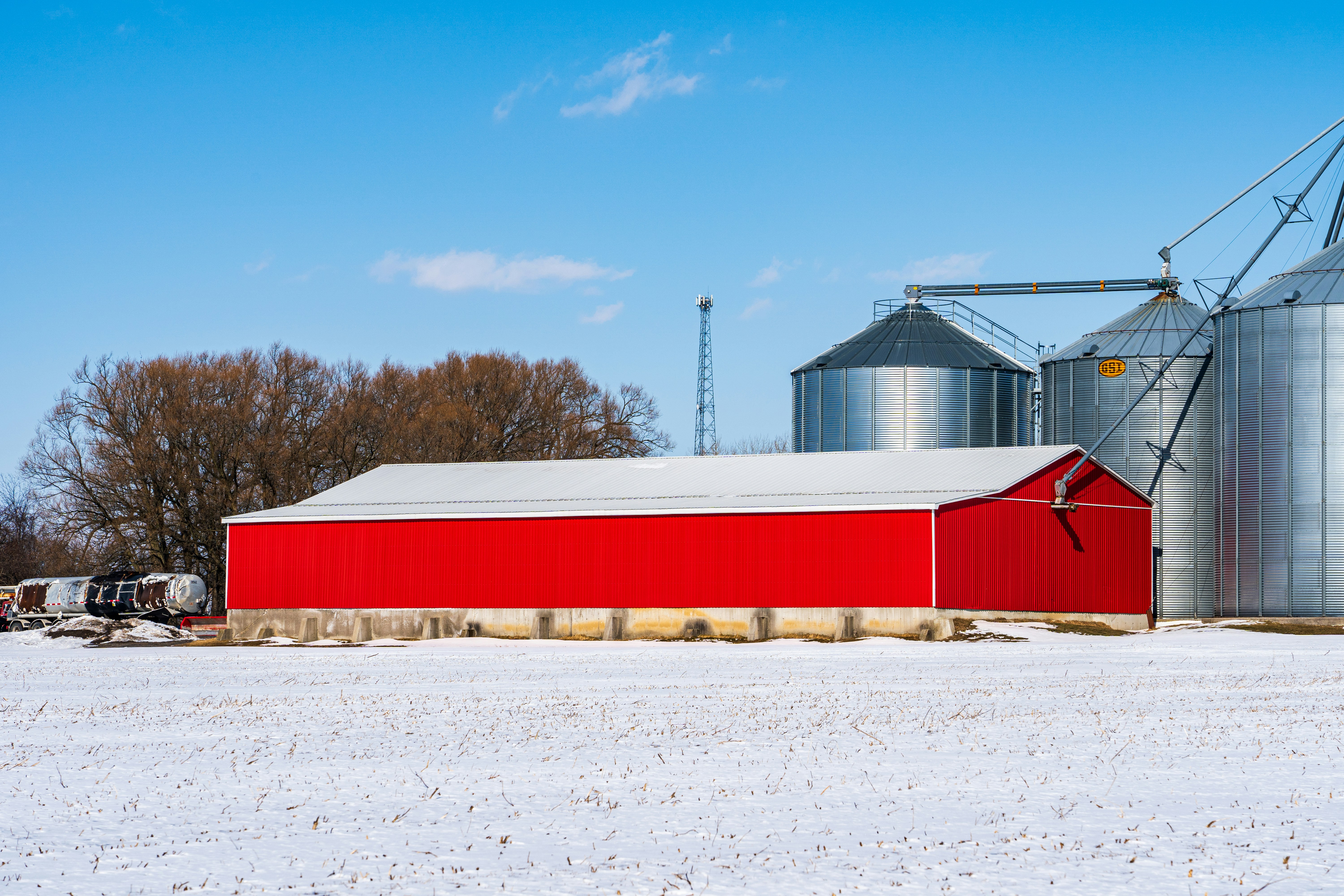 A vibrant red barn stands against a snowy landscape, with silos in the background under a clear blue sky.
