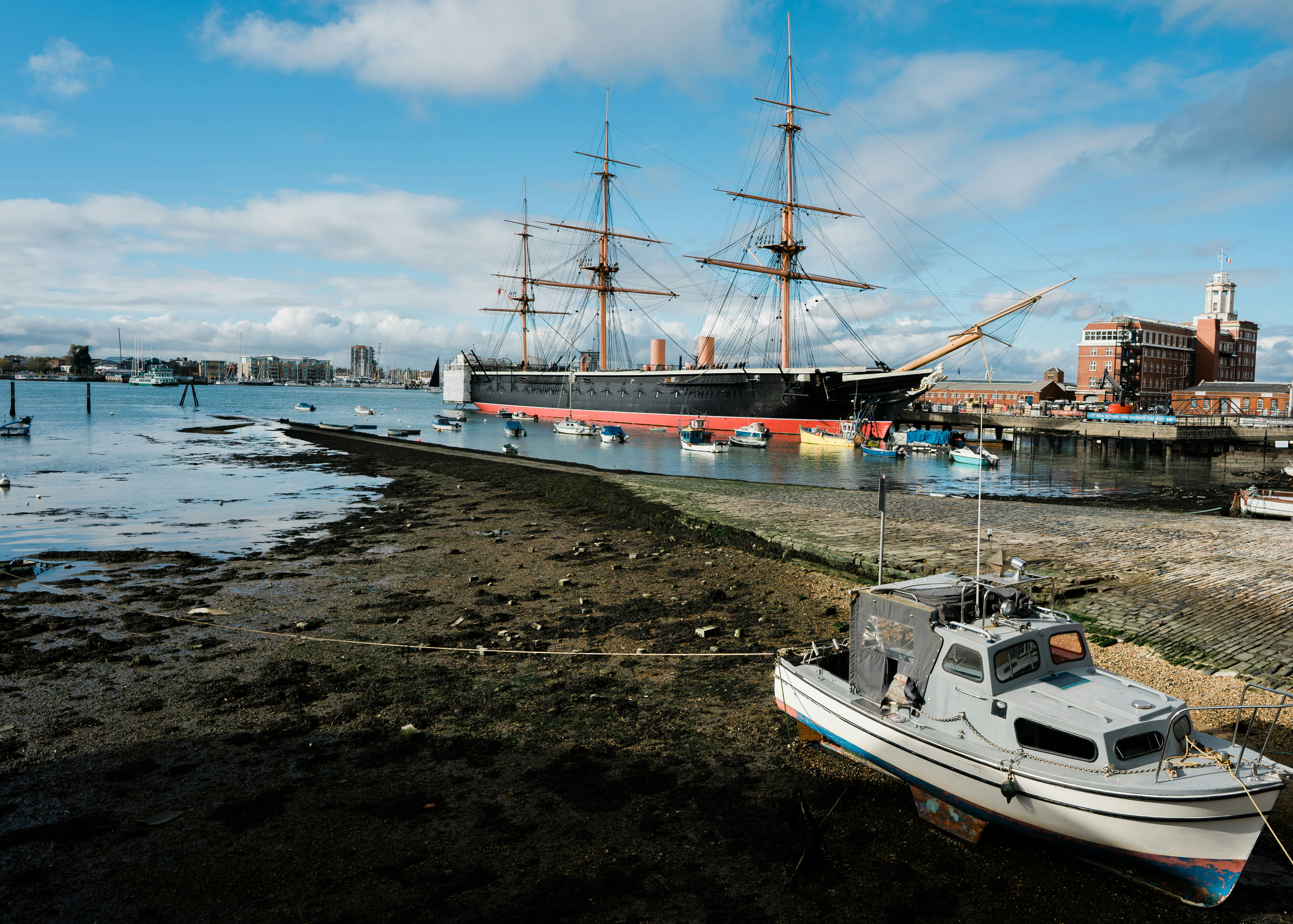 Small boat on a beach with a historic tall ship docked nearby under a bright blue sky.