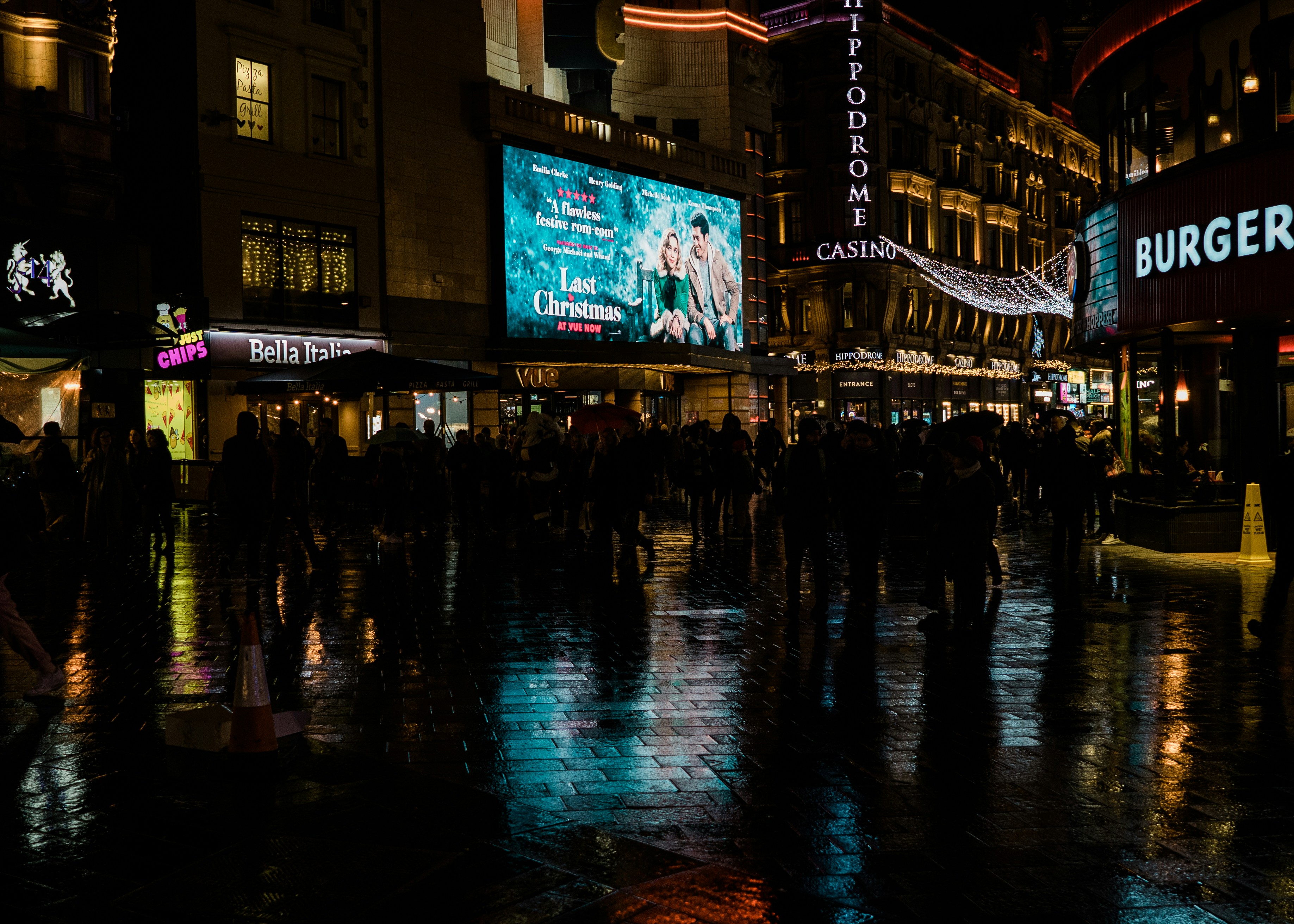 Crowds gather under neon lights in a bustling city square with wet pavement reflecting vibrant colors.