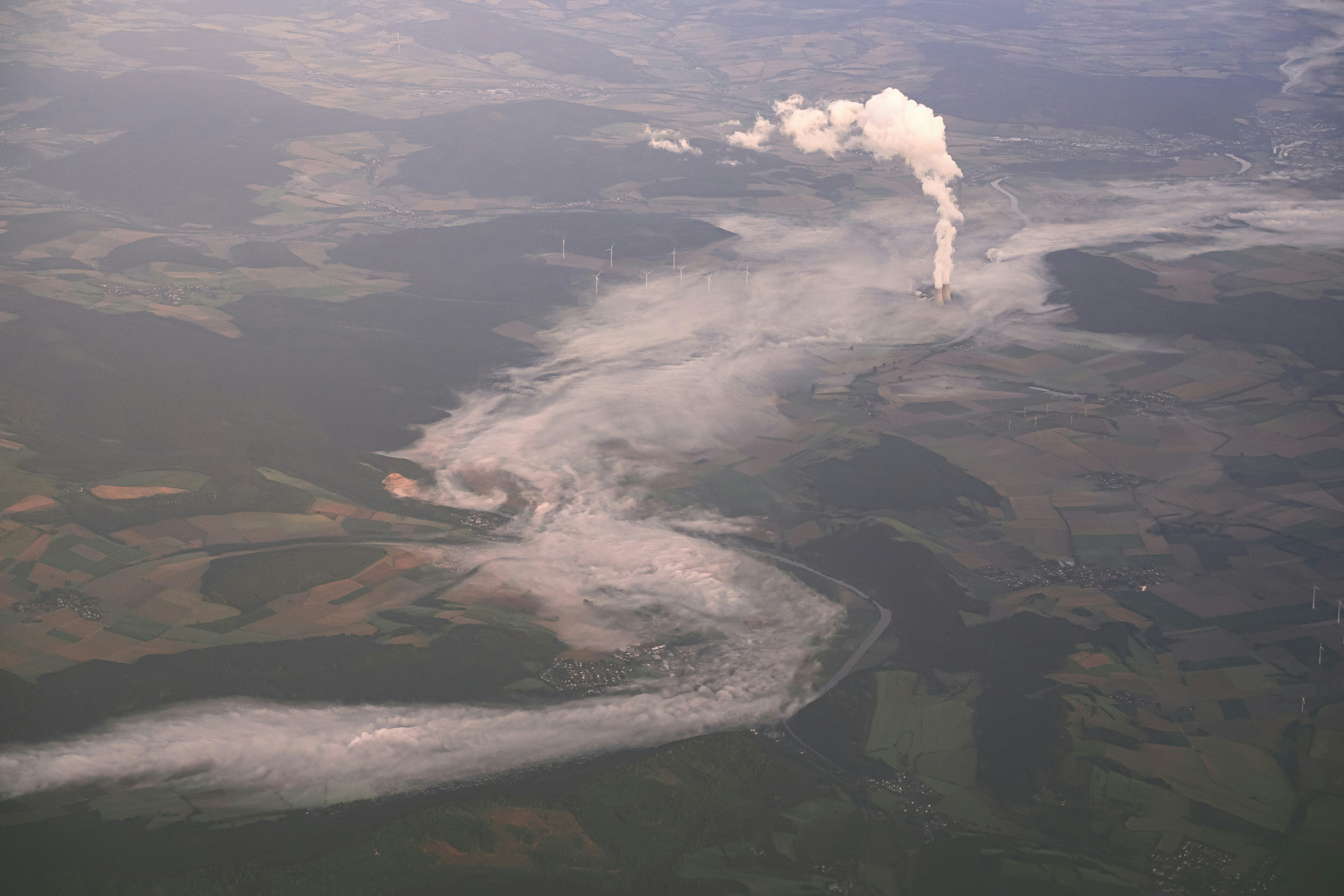 Aerial view showcasing a winding river surrounded by lush landscapes, with smoke rising from a distant industrial site. The scene highlights the contrast between natural beauty and human impact.