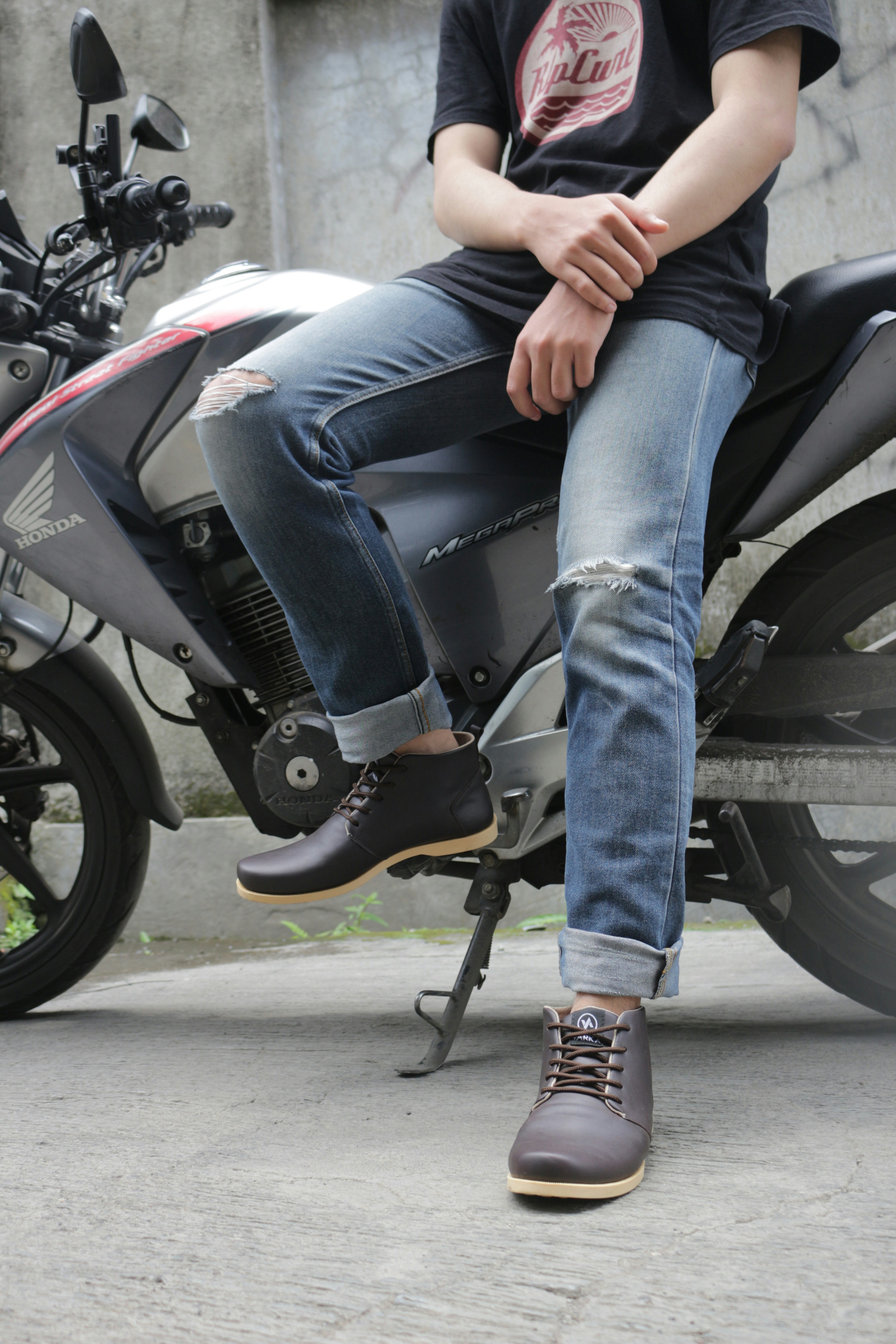A young man casually seated on a motorcycle, showcasing stylish brown shoes and ripped jeans against an urban backdrop.