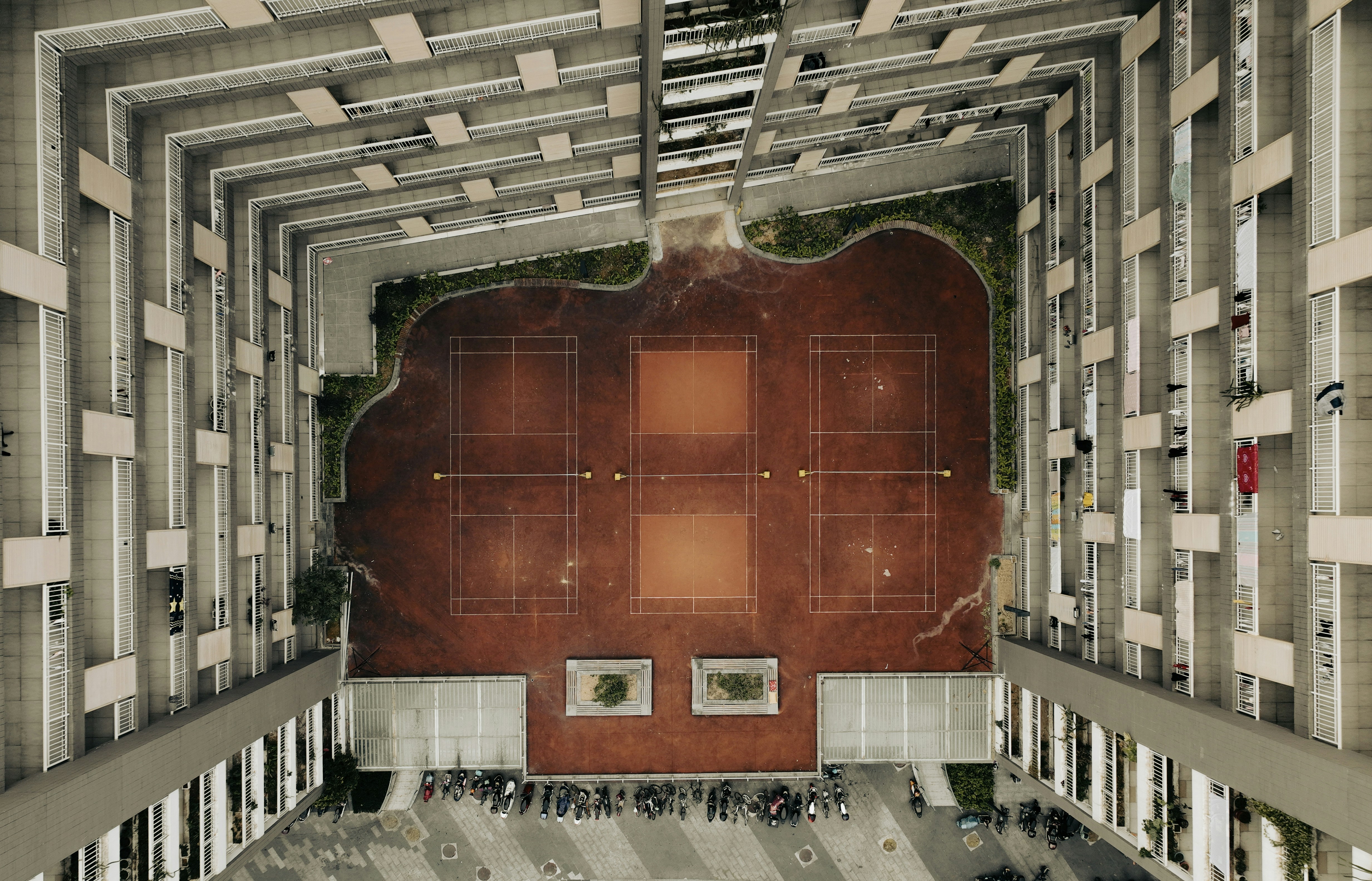 Aerial photograph of a high-rise atrium courtyard featuring four rectangular courts on red clay, surrounded by walkways and balconies. The composition emphasizes symmetry and architectural scale from a top-down perspective.