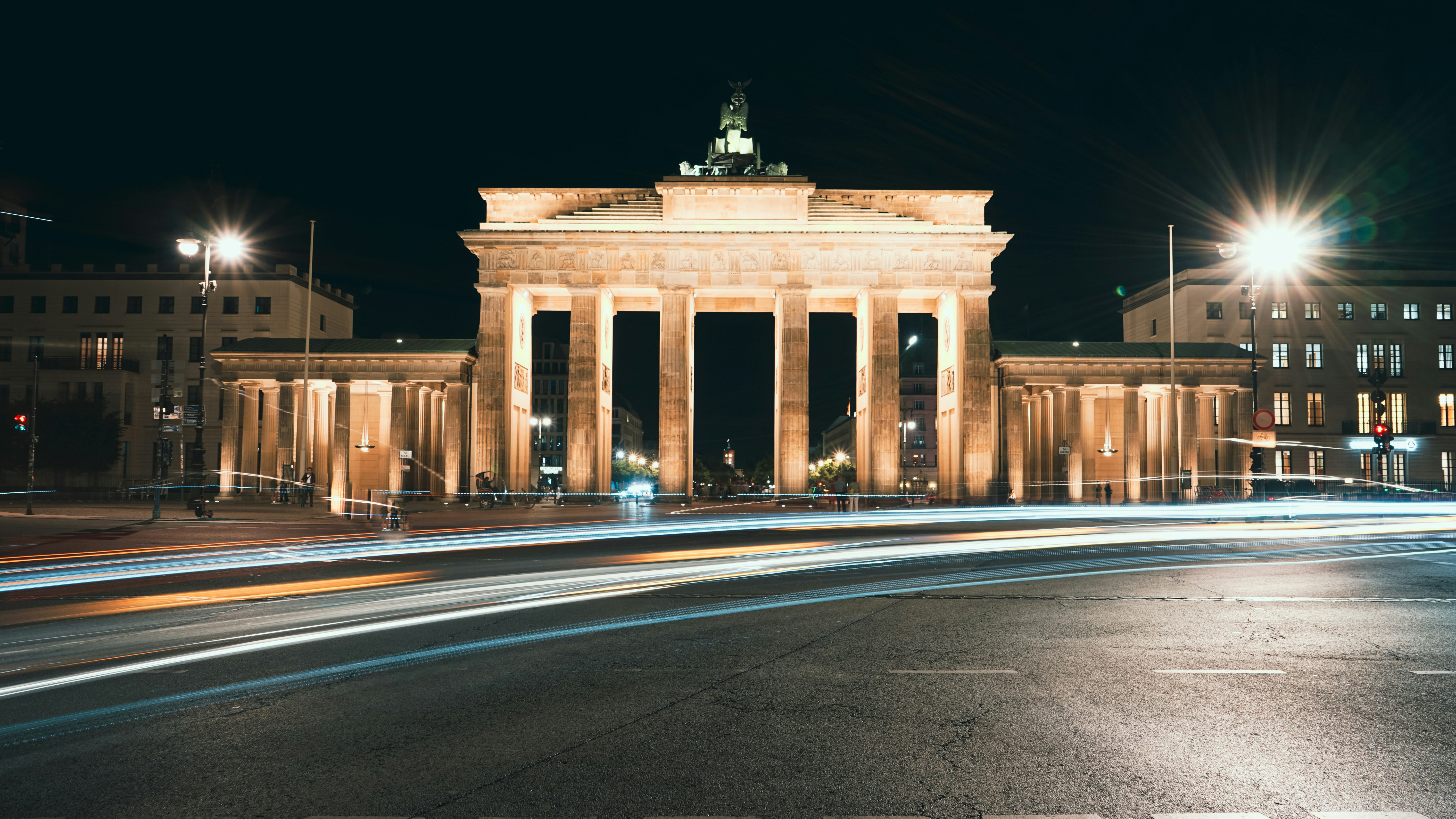 Brandenburg Gate illuminated against the night sky, surrounded by light trails from passing vehicles. A symbol of history and unity.