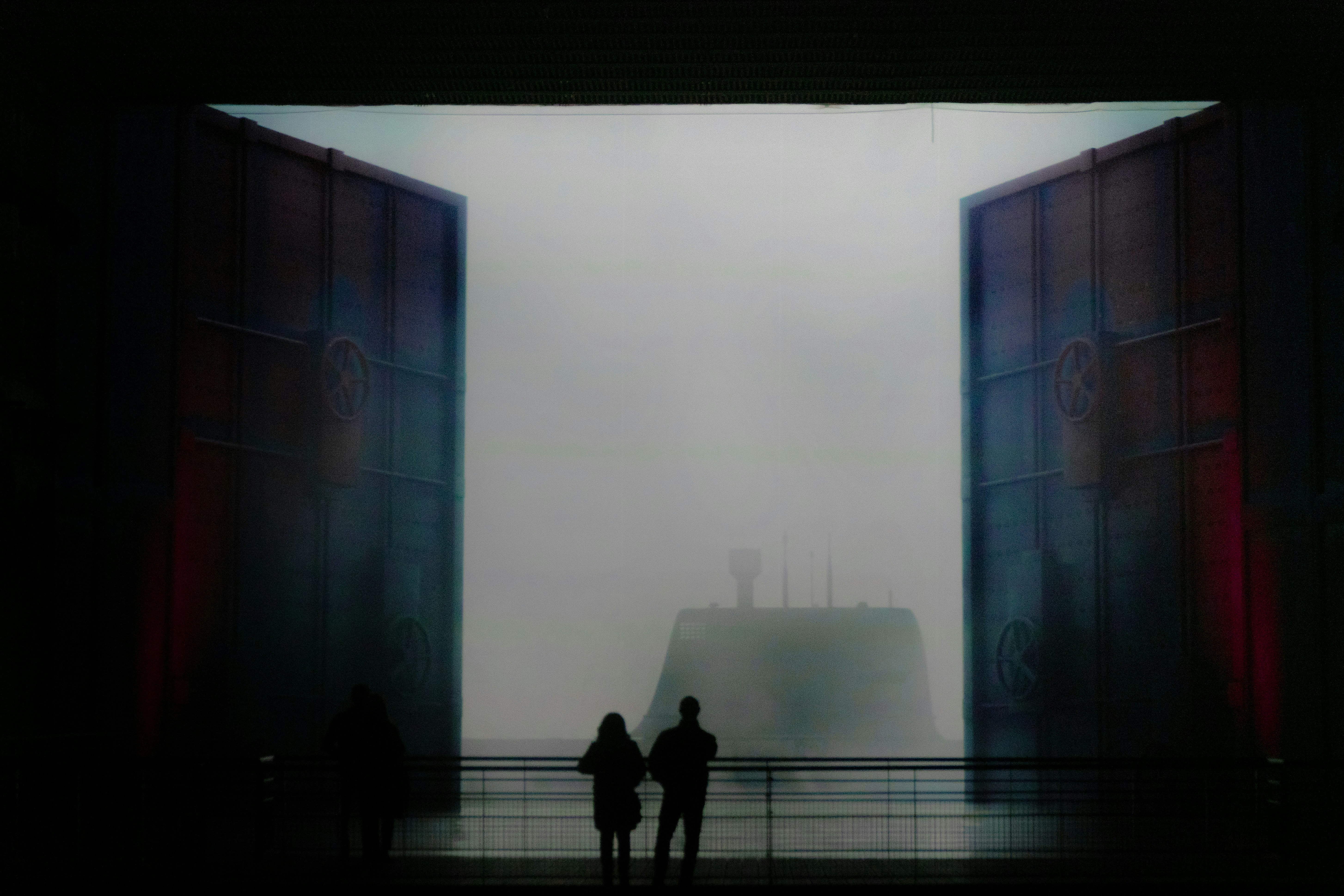 Silhouetted figures stand before large, partially open gates shrouded in fog, hinting at an enigmatic scene beyond.