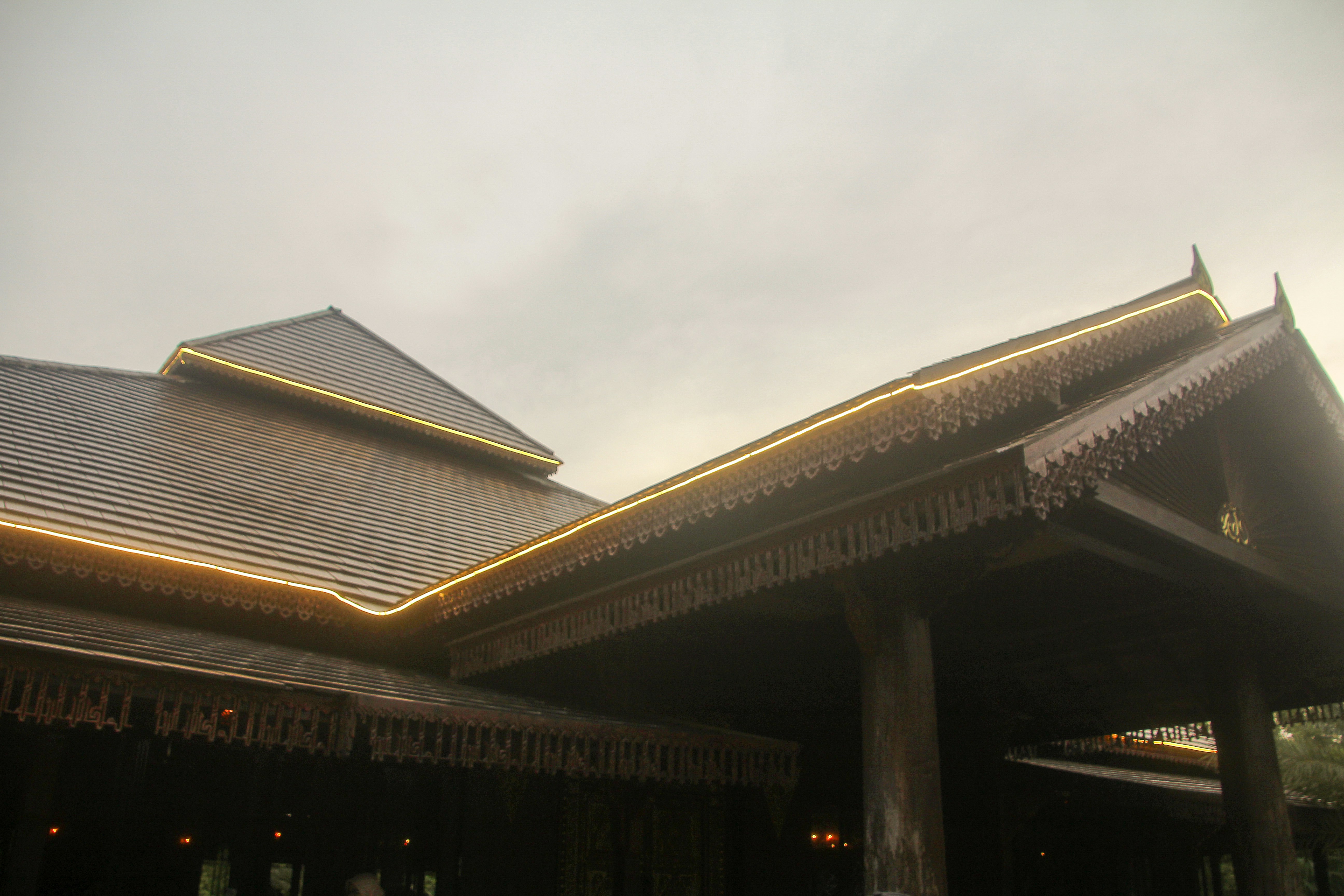 Elegant wooden architecture with intricate eaves under a cloudy sky, showcasing traditional design elements. The warm glow of lights highlights the craftsmanship.