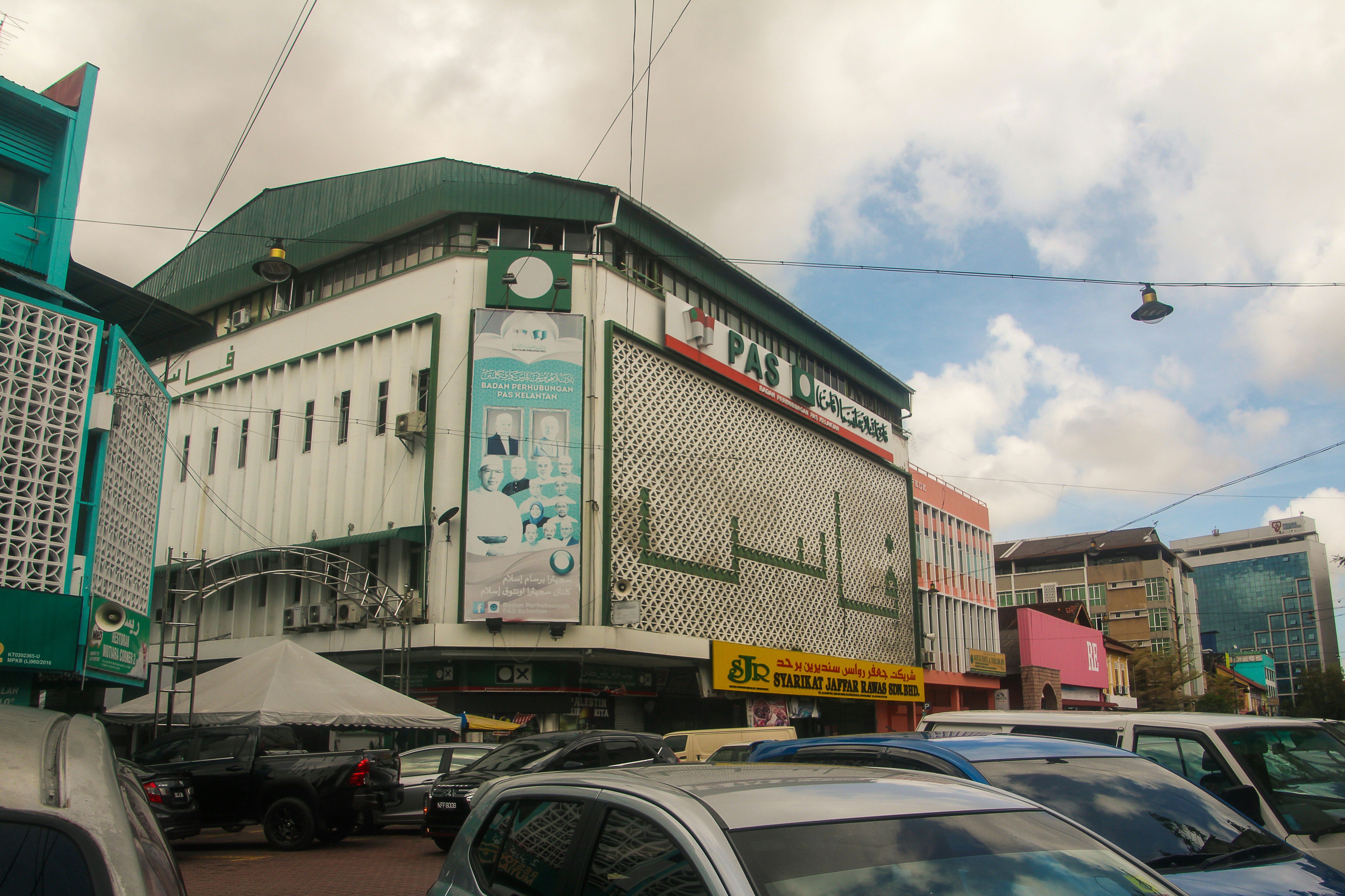 Busy street scene with parked cars and multi-story buildings under a partly cloudy sky.