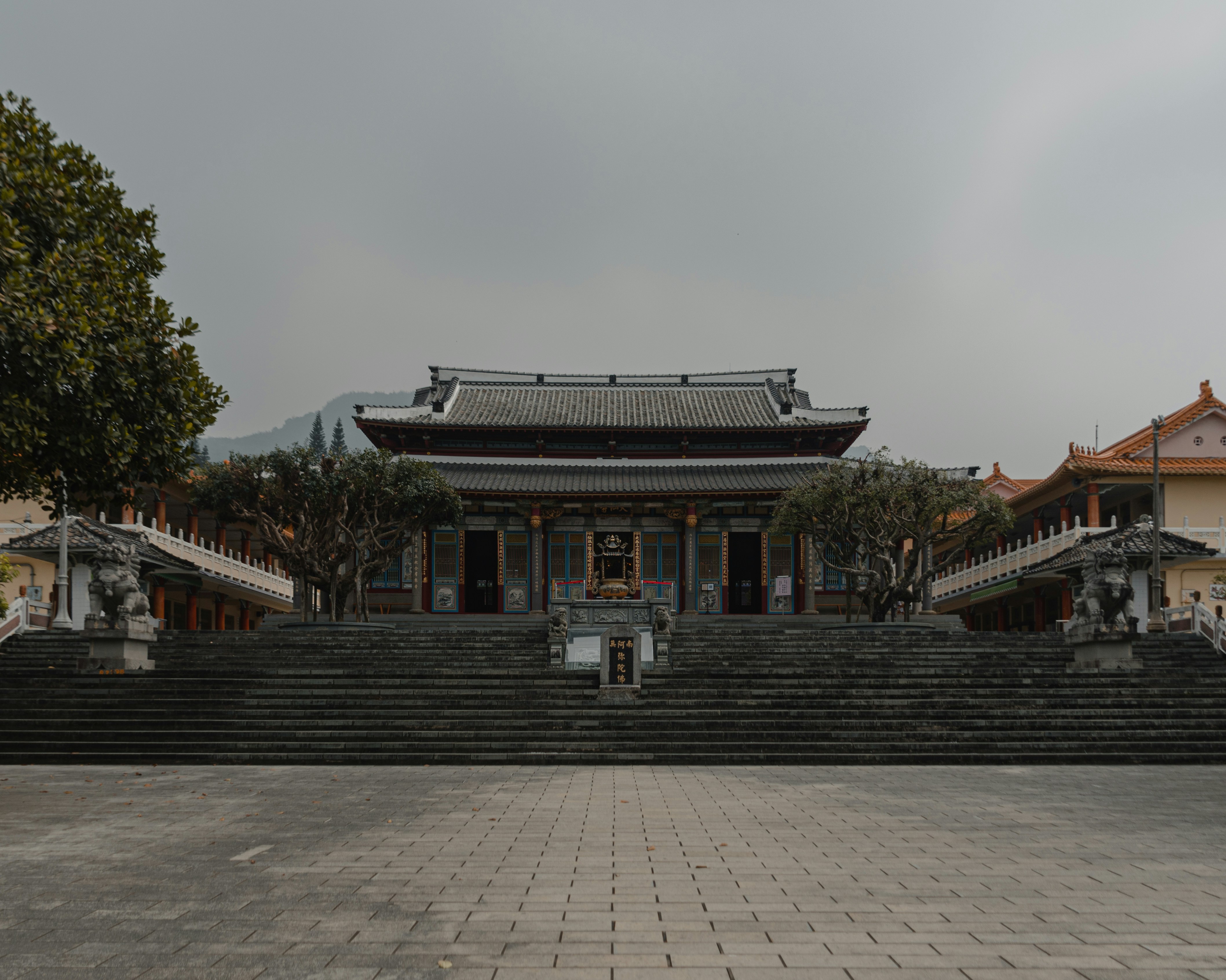A grand temple with intricate architecture, flanked by stone lions and surrounded by trees, set against a muted sky.