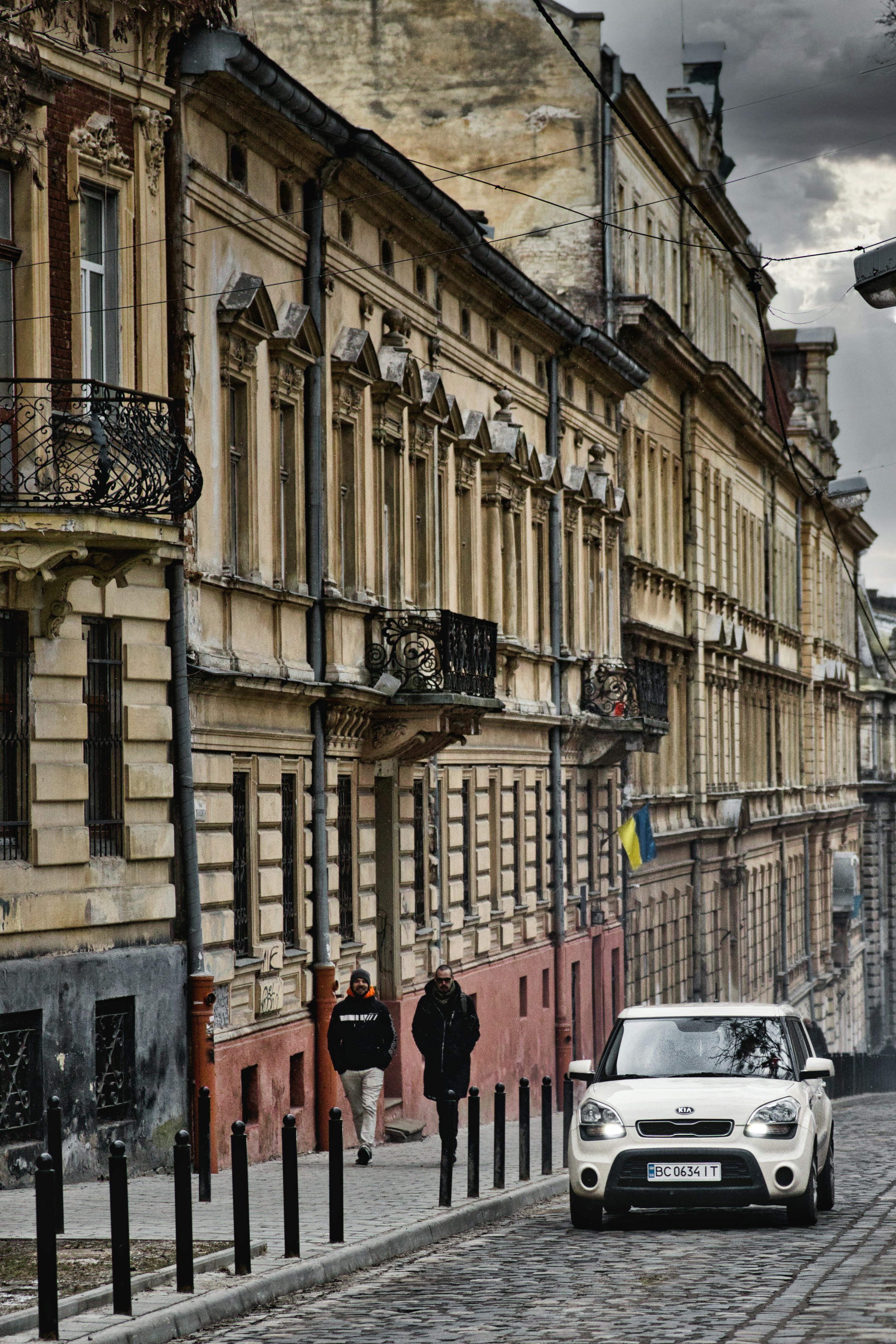 White car navigating a cobblestone street lined with historic buildings, as two pedestrians stroll by. A Ukrainian flag flutters in the background.
