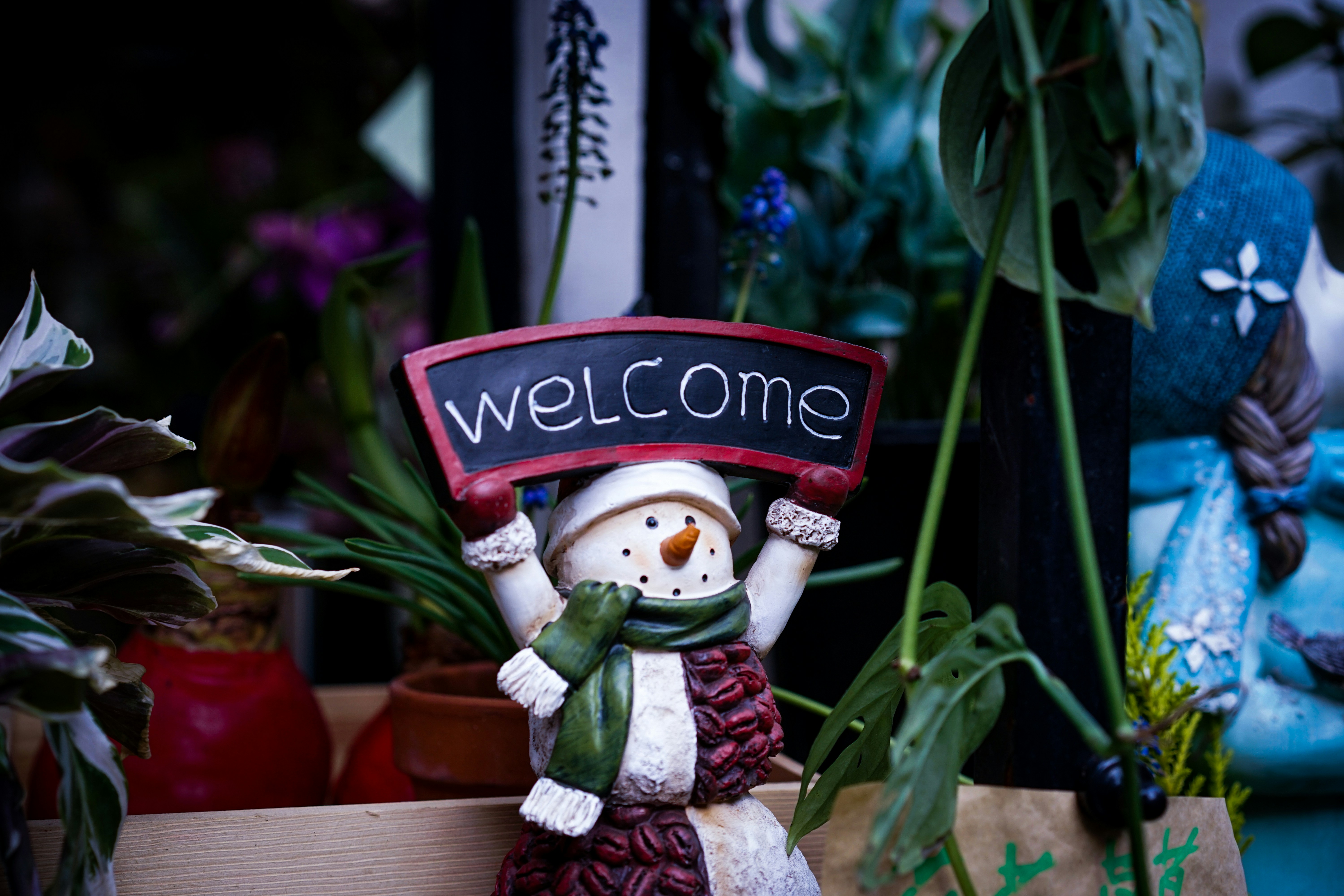 A cheerful snowman holding a chalkboard sign that reads 'welcome,' surrounded by vibrant plants and decorations.