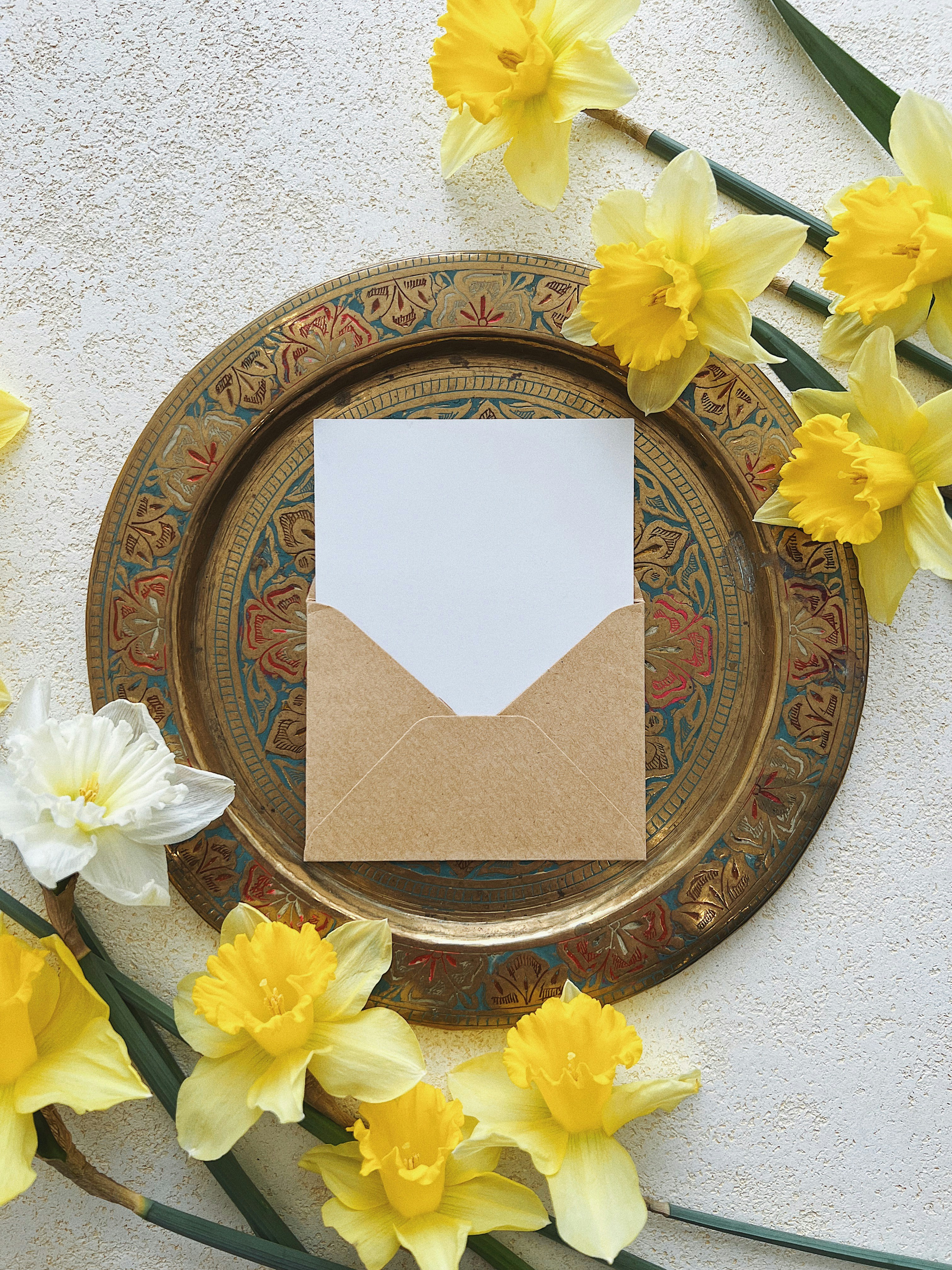 Flat lay photograph of a brown envelope with a white card on a decorative brass plate, surrounded by bright yellow daffodils.