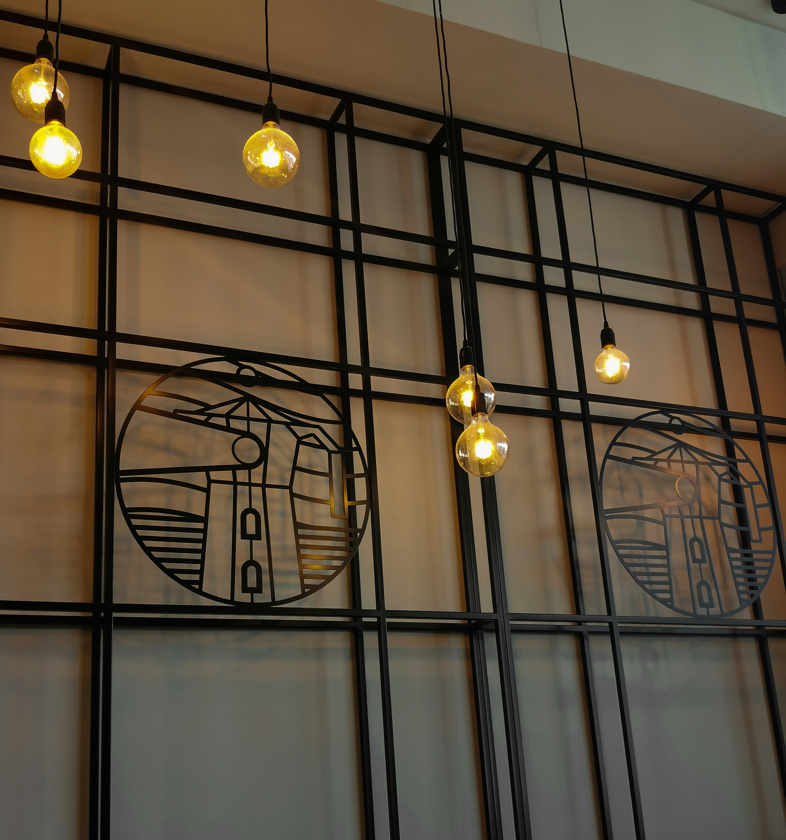 Warm Edison bulbs hang over a black metal grid wall with circular emblem panels, creating a moody, geometric backdrop.