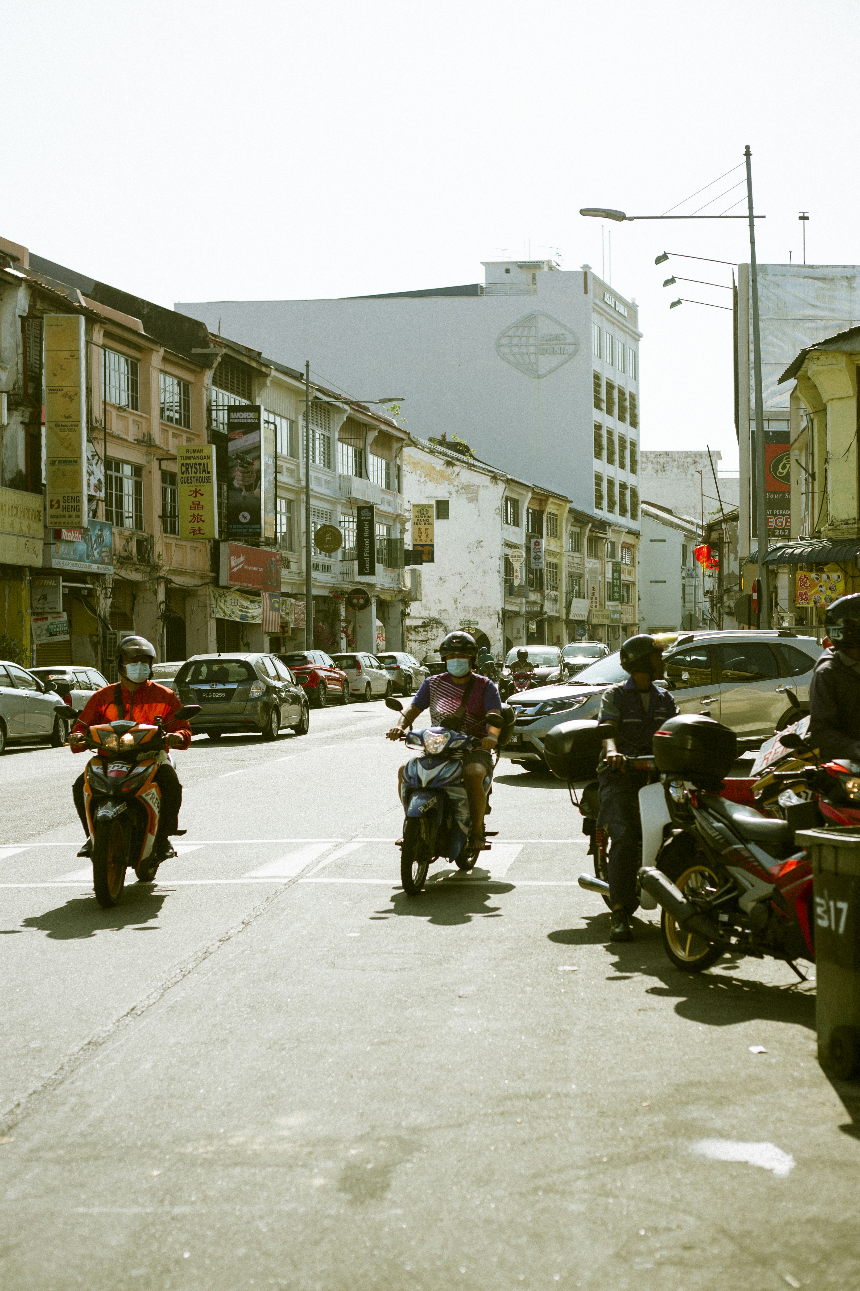 Motorcyclists navigate a busy urban street lined with colorful buildings under bright sunlight.