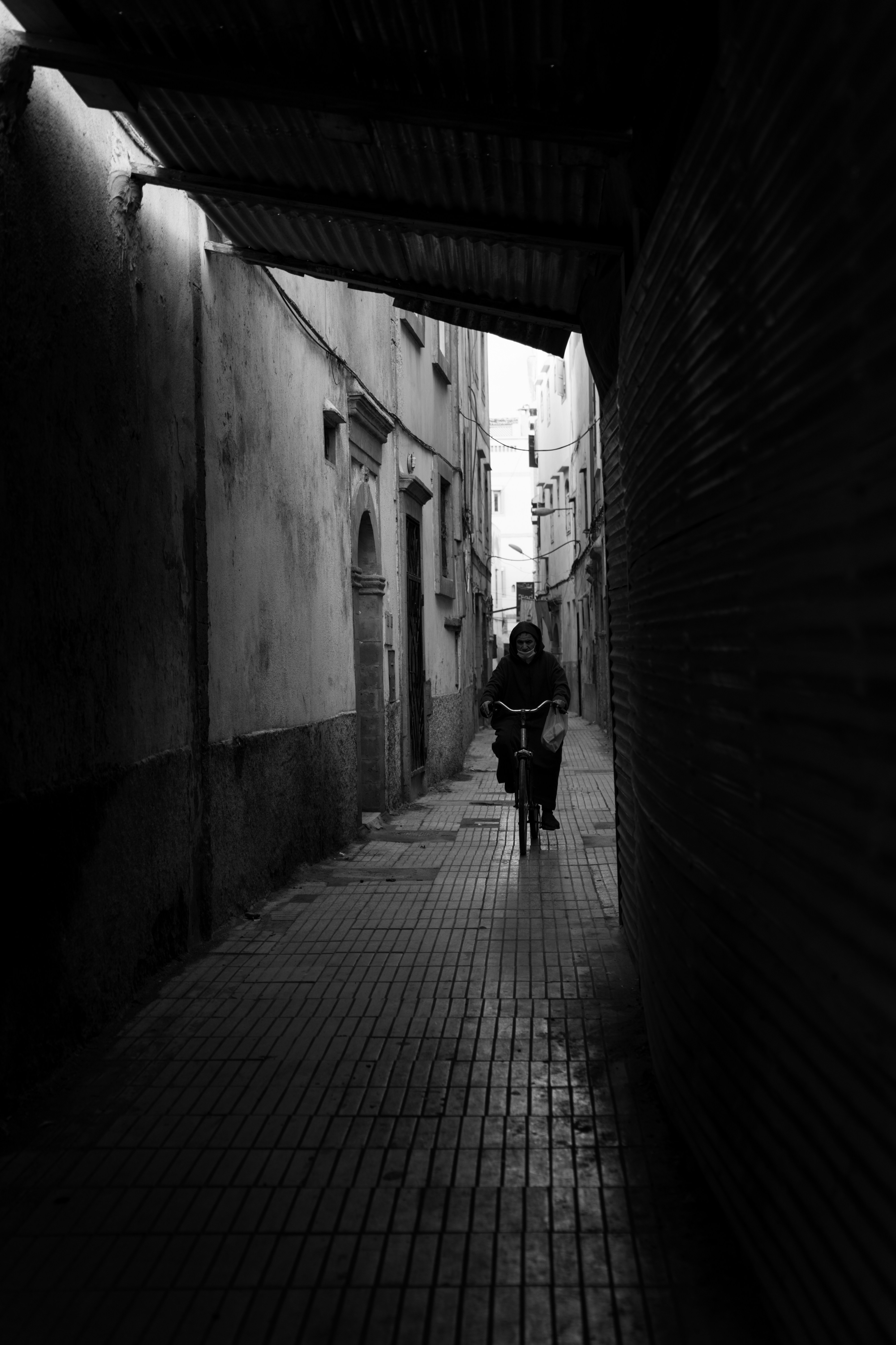 A cyclist navigates a narrow, dimly lit alleyway, flanked by weathered walls and a textured pavement. The scene evokes a sense of solitude and exploration.