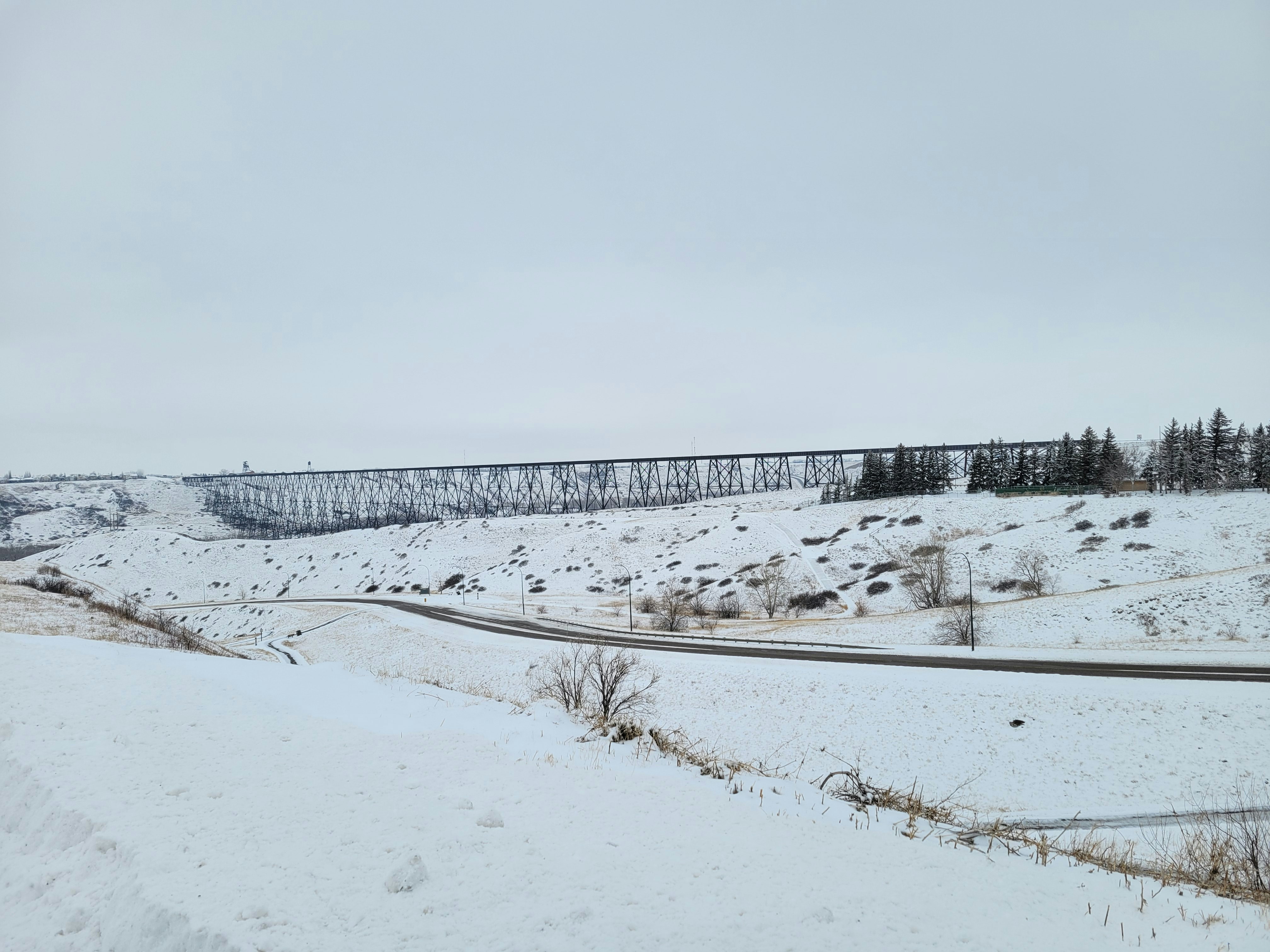 Snow-covered landscape with a distant railway bridge under a cloudy sky.