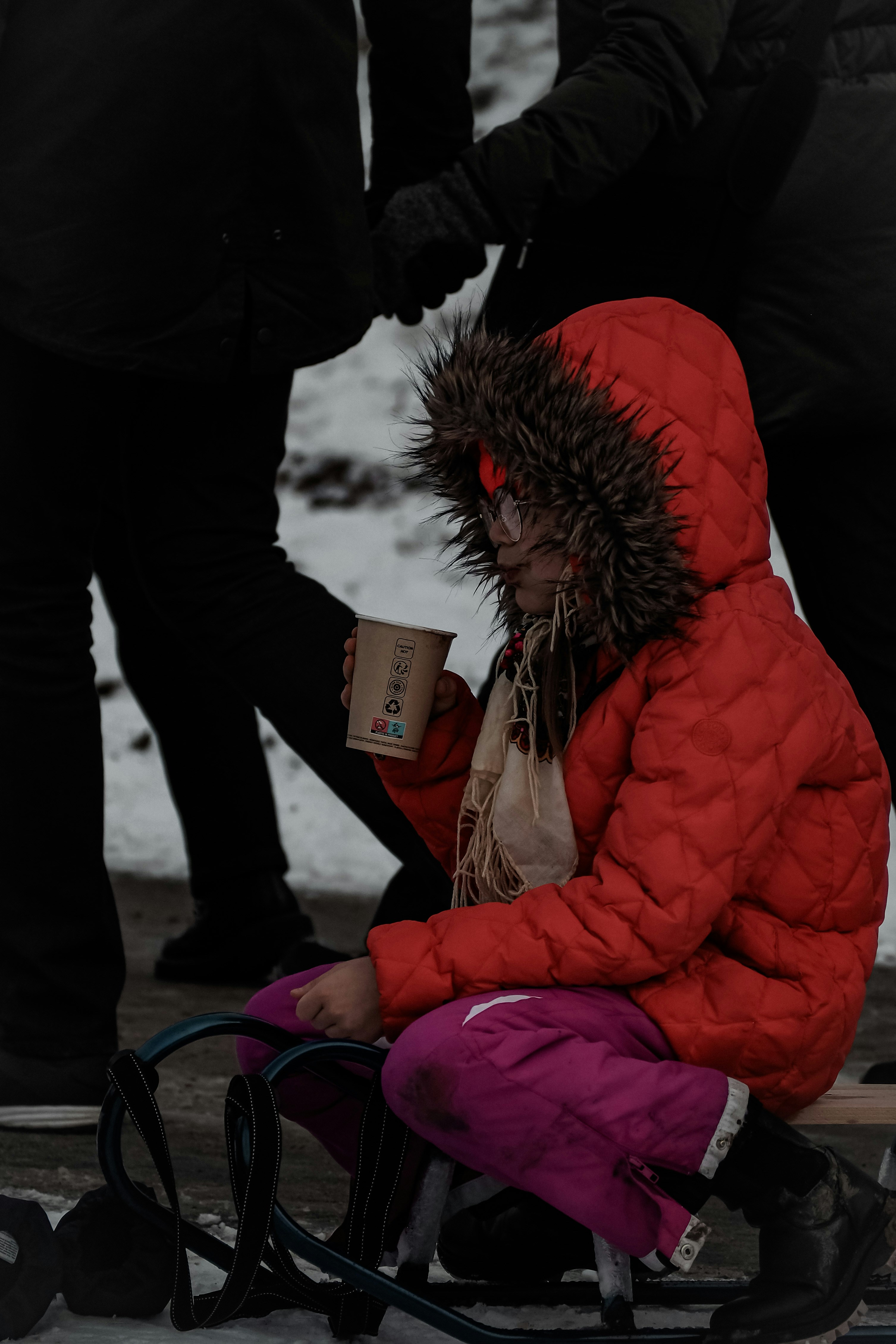 Child in a red coat and pink pants sits on the ground, sipping a drink while surrounded by adults in winter attire.