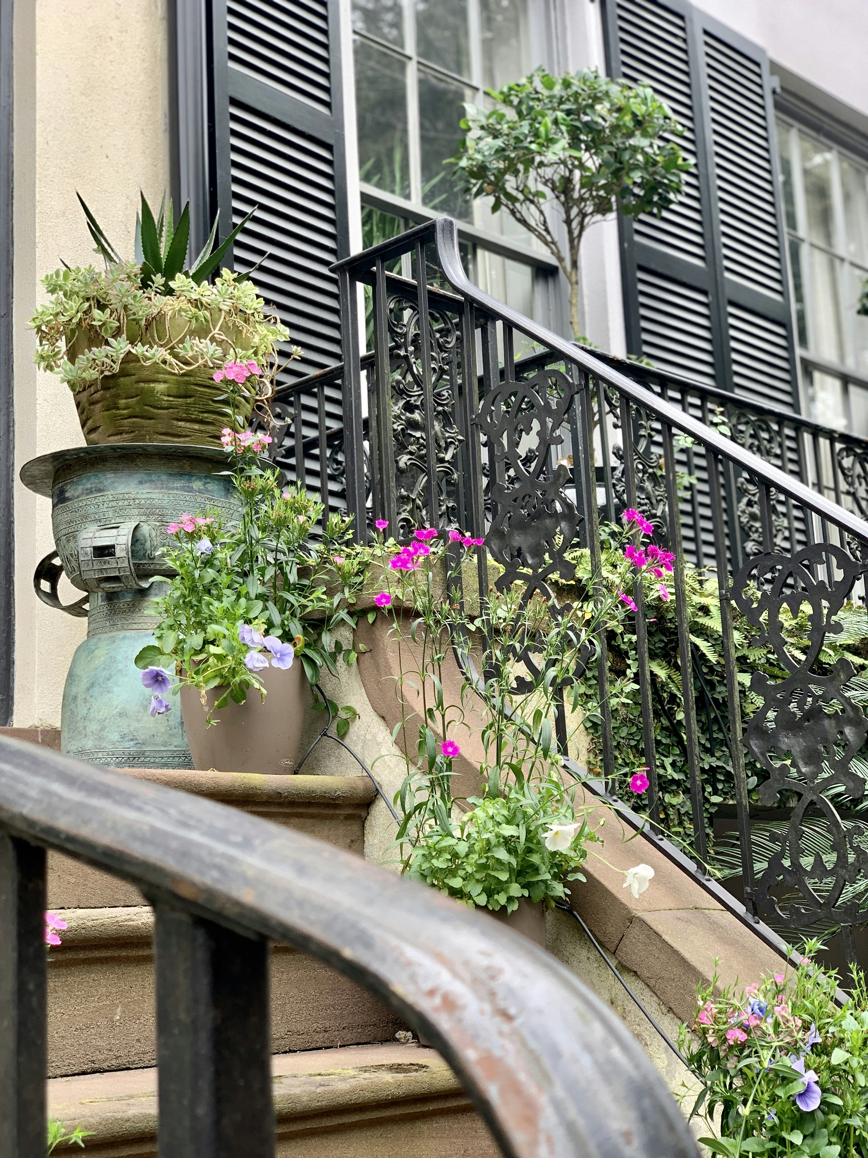 Vibrant flowers cascade down stone steps, framed by elegant ironwork and lush greenery. A vintage planter adds character to the inviting entrance.