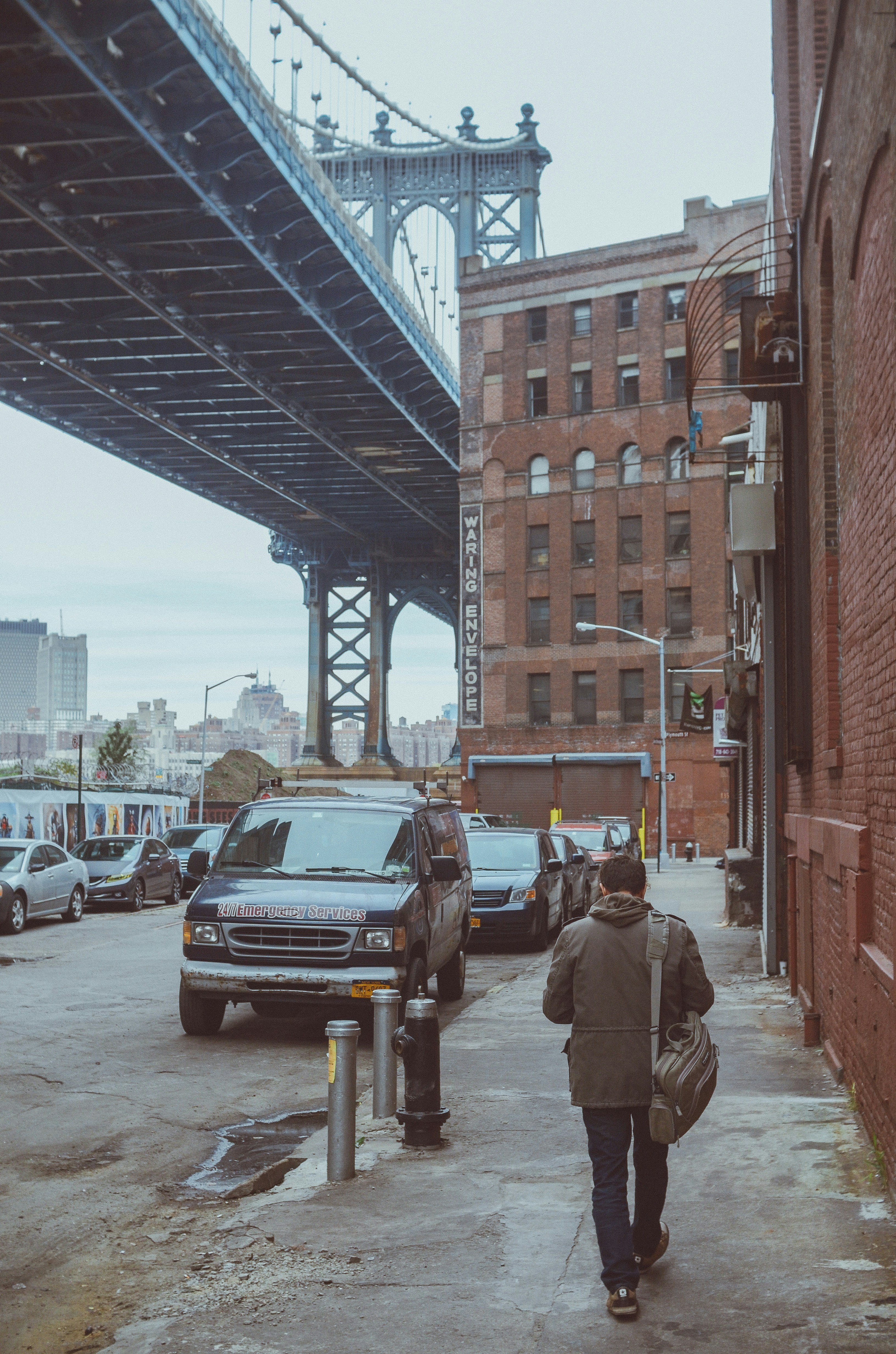 A lone pedestrian walks along a city street beneath a towering bridge, flanked by vintage brick buildings and parked vehicles.