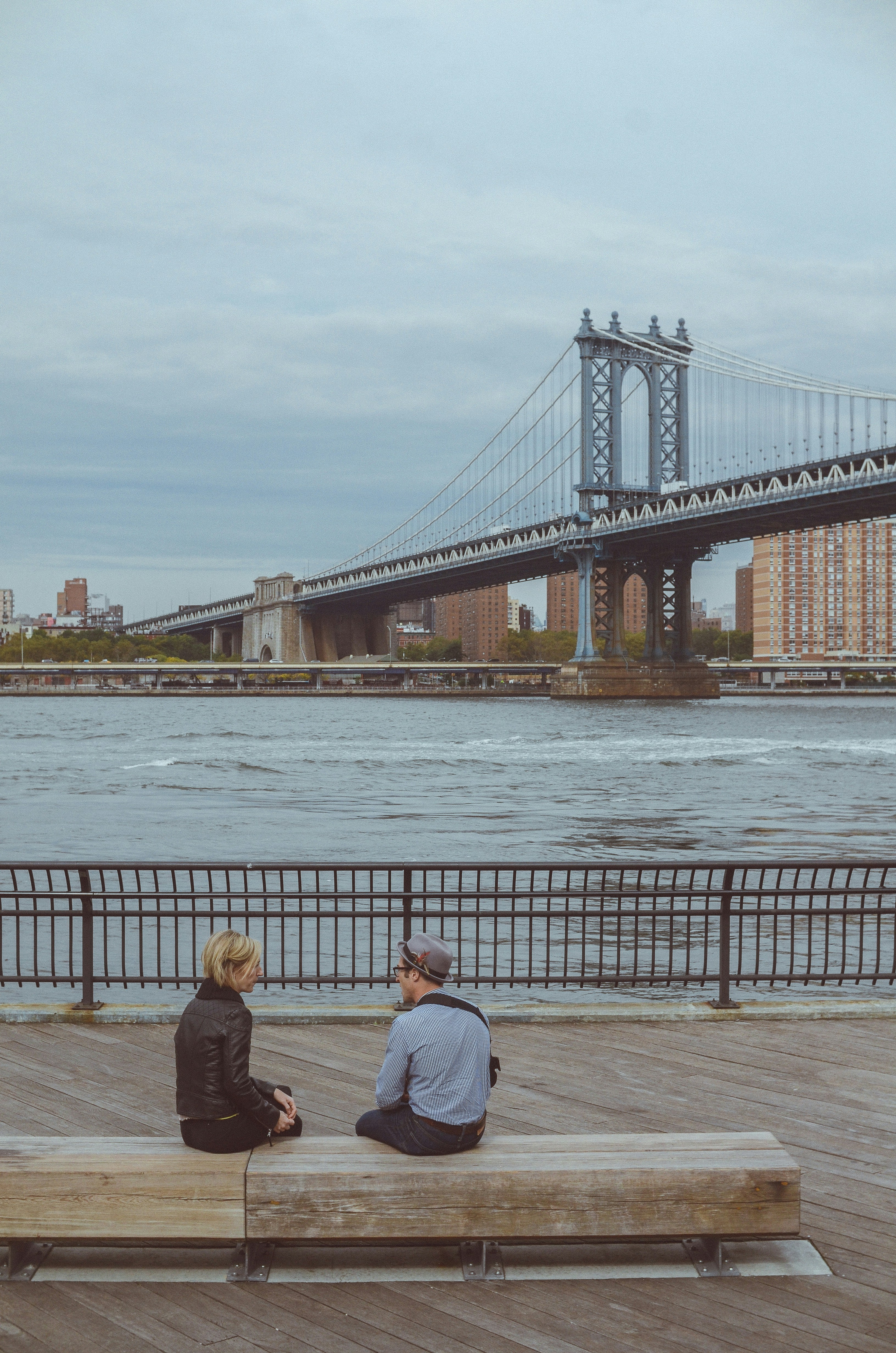 Two individuals seated on a bench, engaged in conversation, with the Manhattan Bridge and city skyline in the background. The scene captures a serene urban moment.